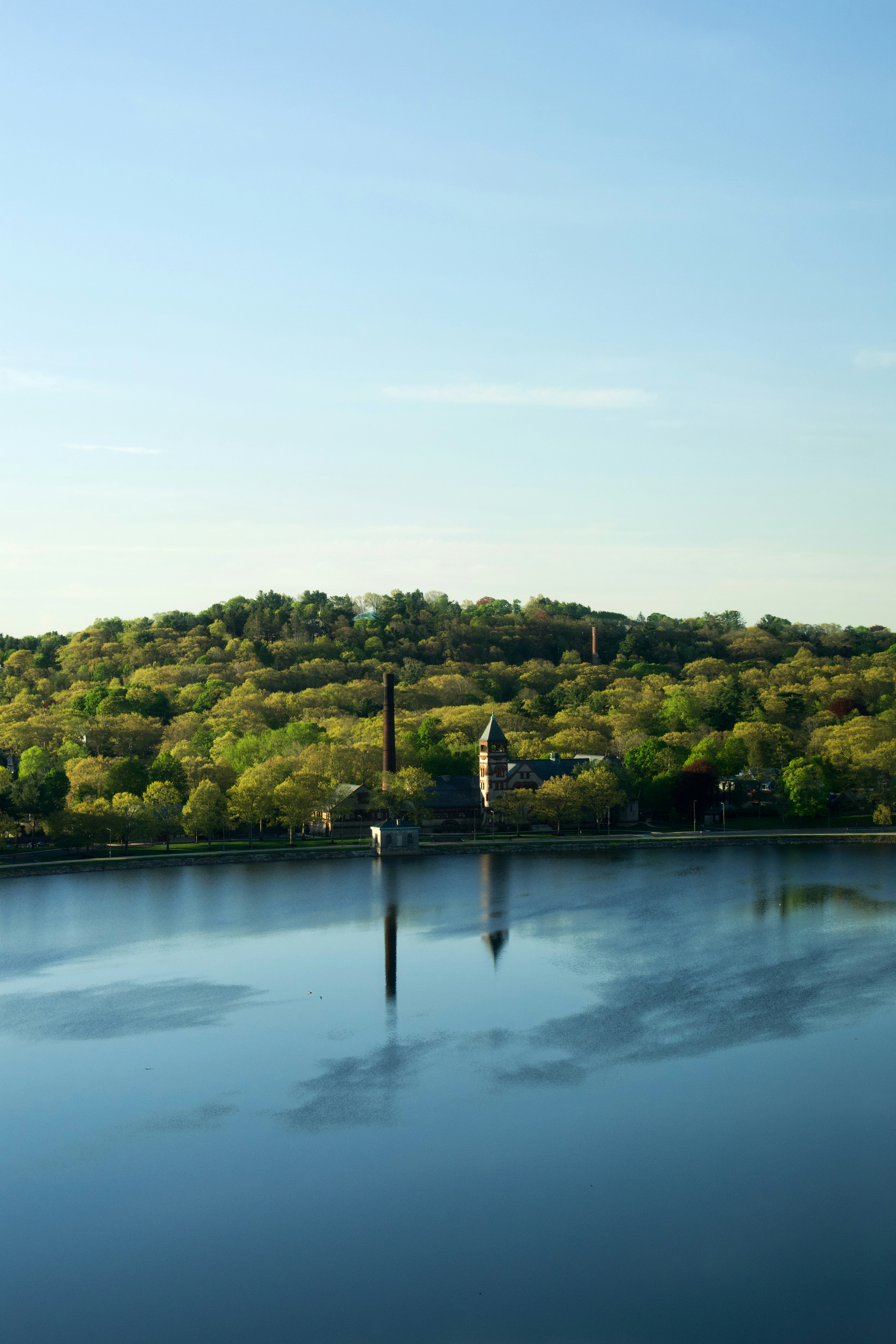 Lush green hills reflect in a calm lake under a clear sky, with an old building and smokestacks visible along the shore.