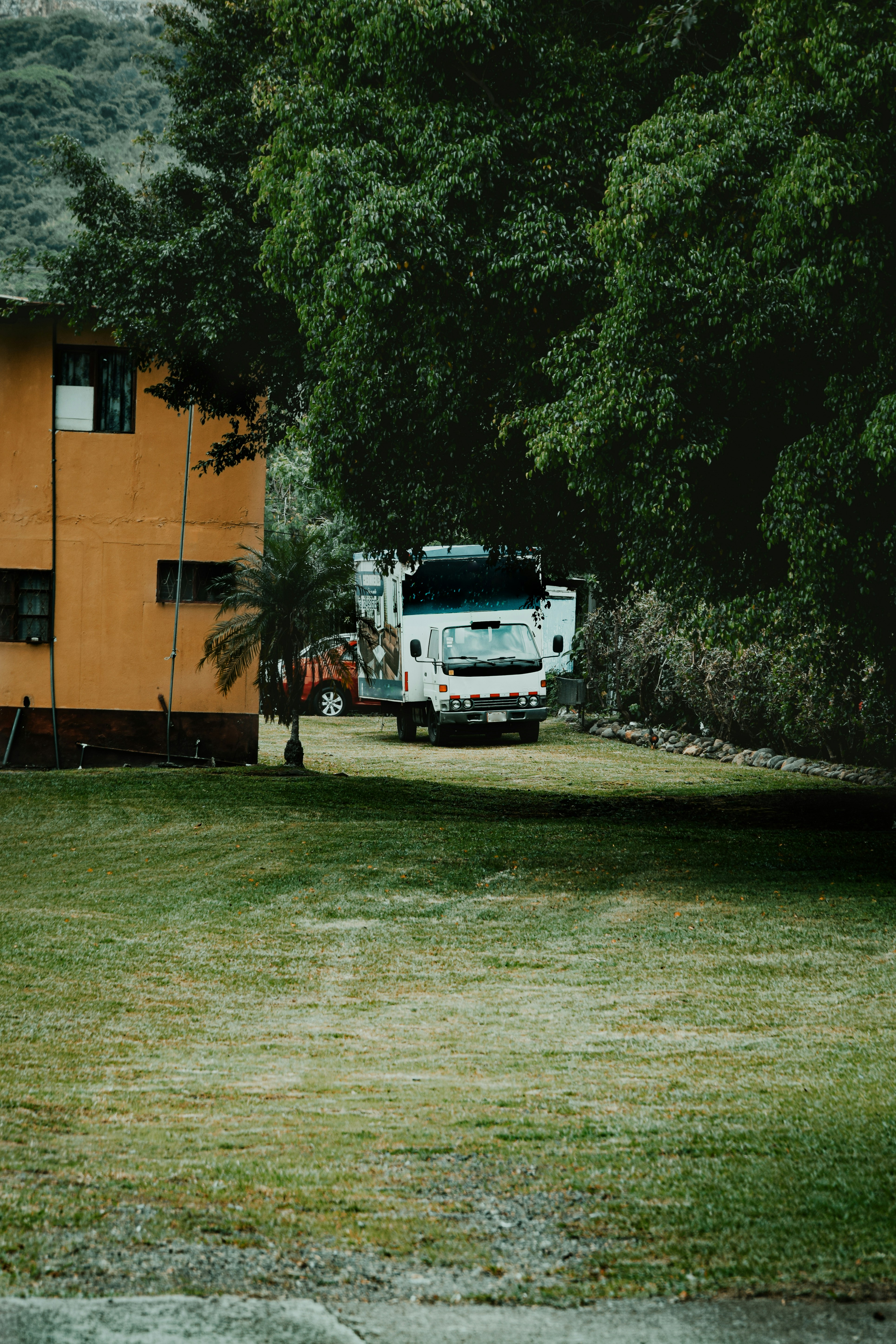 A white vehicle parked near a vibrant orange building, surrounded by lush greenery and distant mountains.