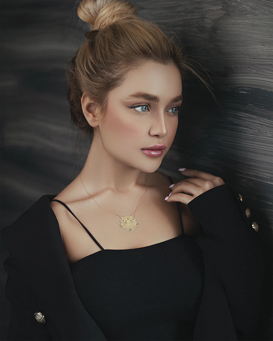 Elegant studio portrait of a woman in a black off-shoulder outfit, with a high bun, gold necklace, and soft side profile. Dark brush-stroked background adds contrast and mood.
