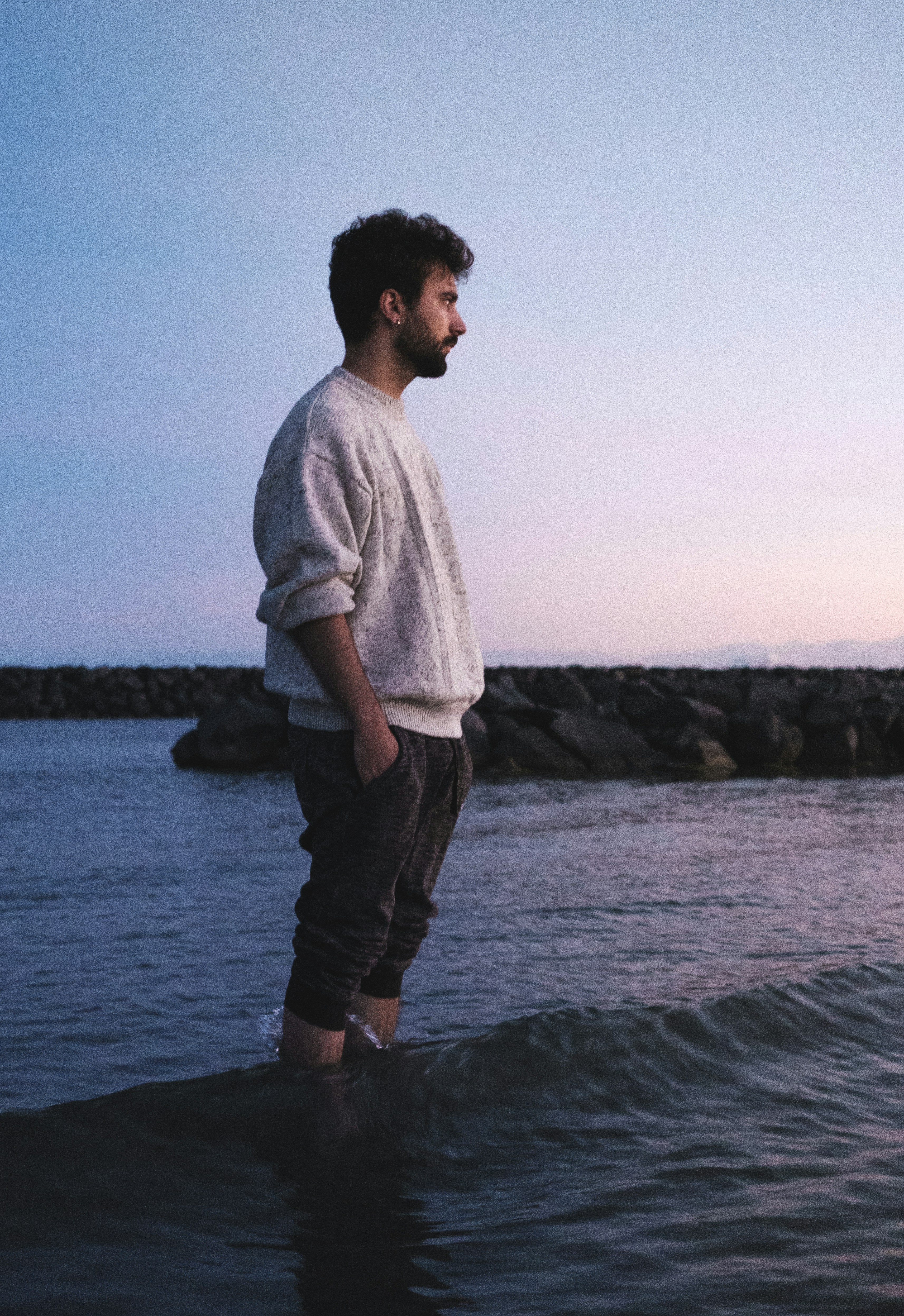 Bearded man walking on a beach, waves crashing on his legs. Sunset, blue and pink mood