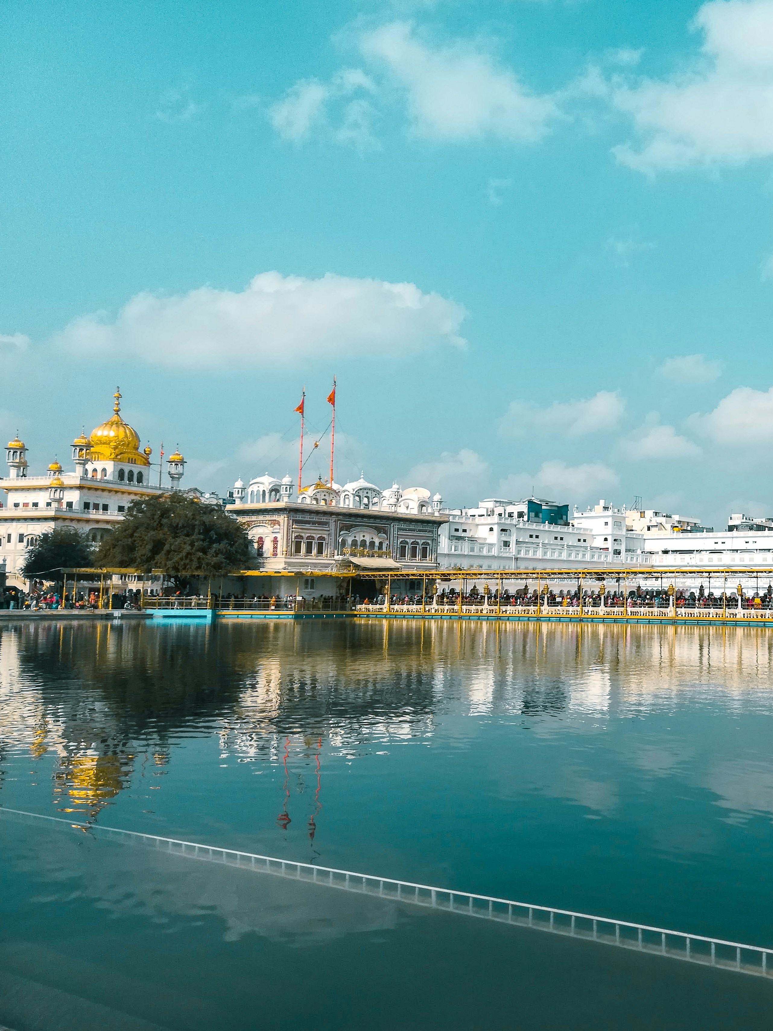 Golden temple reflecting in tranquil waters under a clear blue sky, surrounded by visitors and lush greenery.