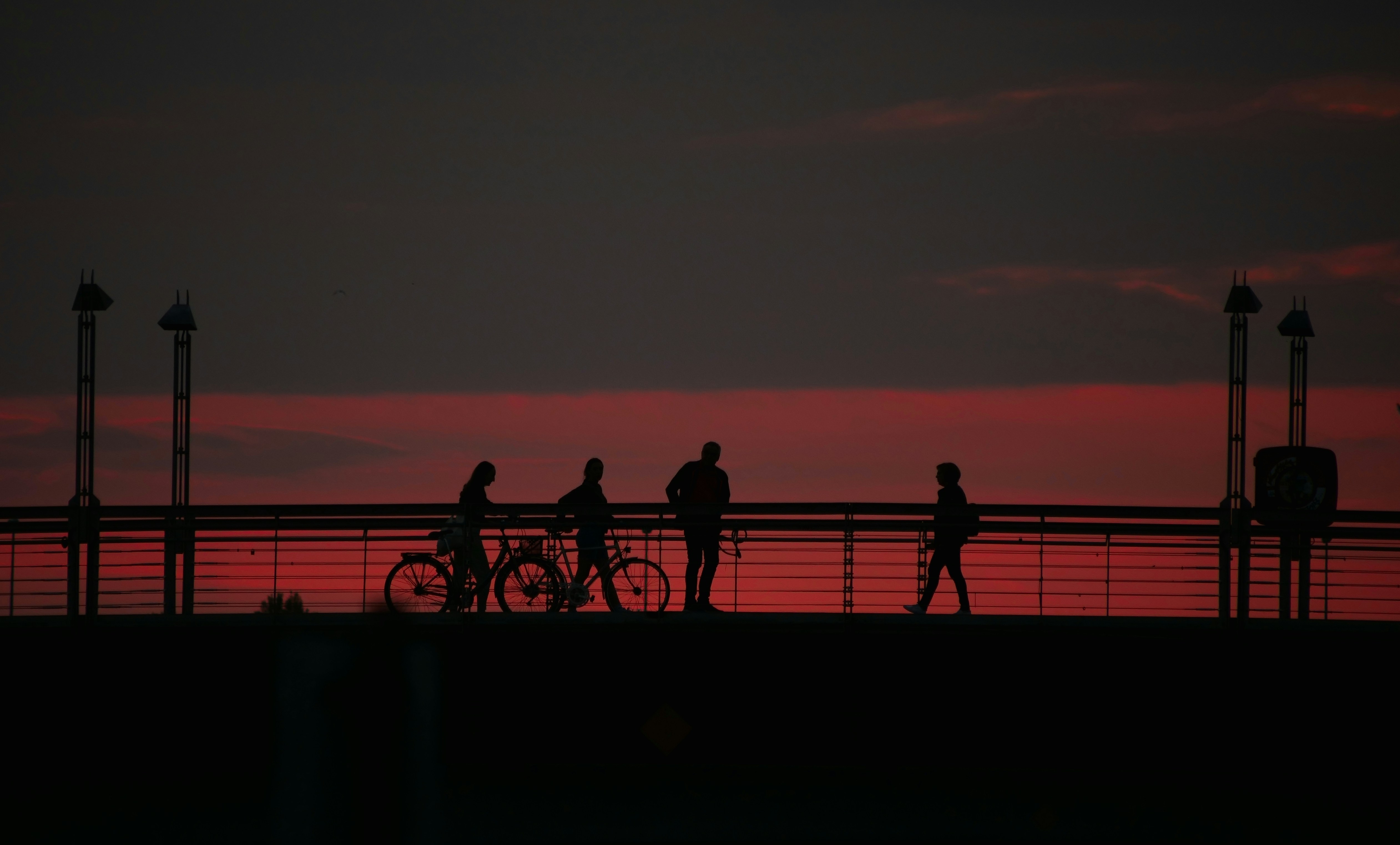 Silhouetted figures and a bicycle on a bridge against a deep red sunset sky.