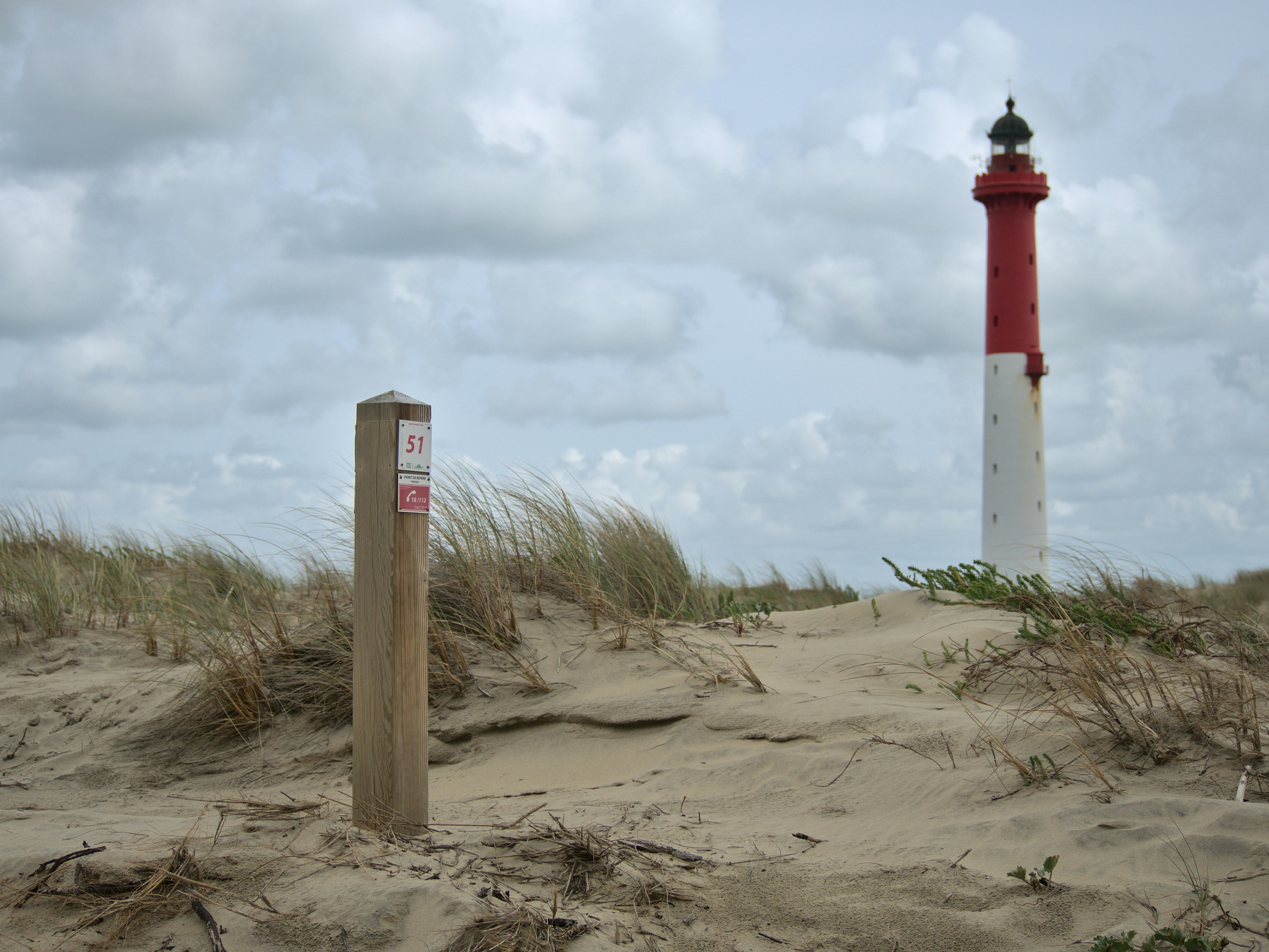 Weathered post marking a sandy dune with a distant lighthouse in the background. The scene captures the interplay between natural elements and man-made structures.