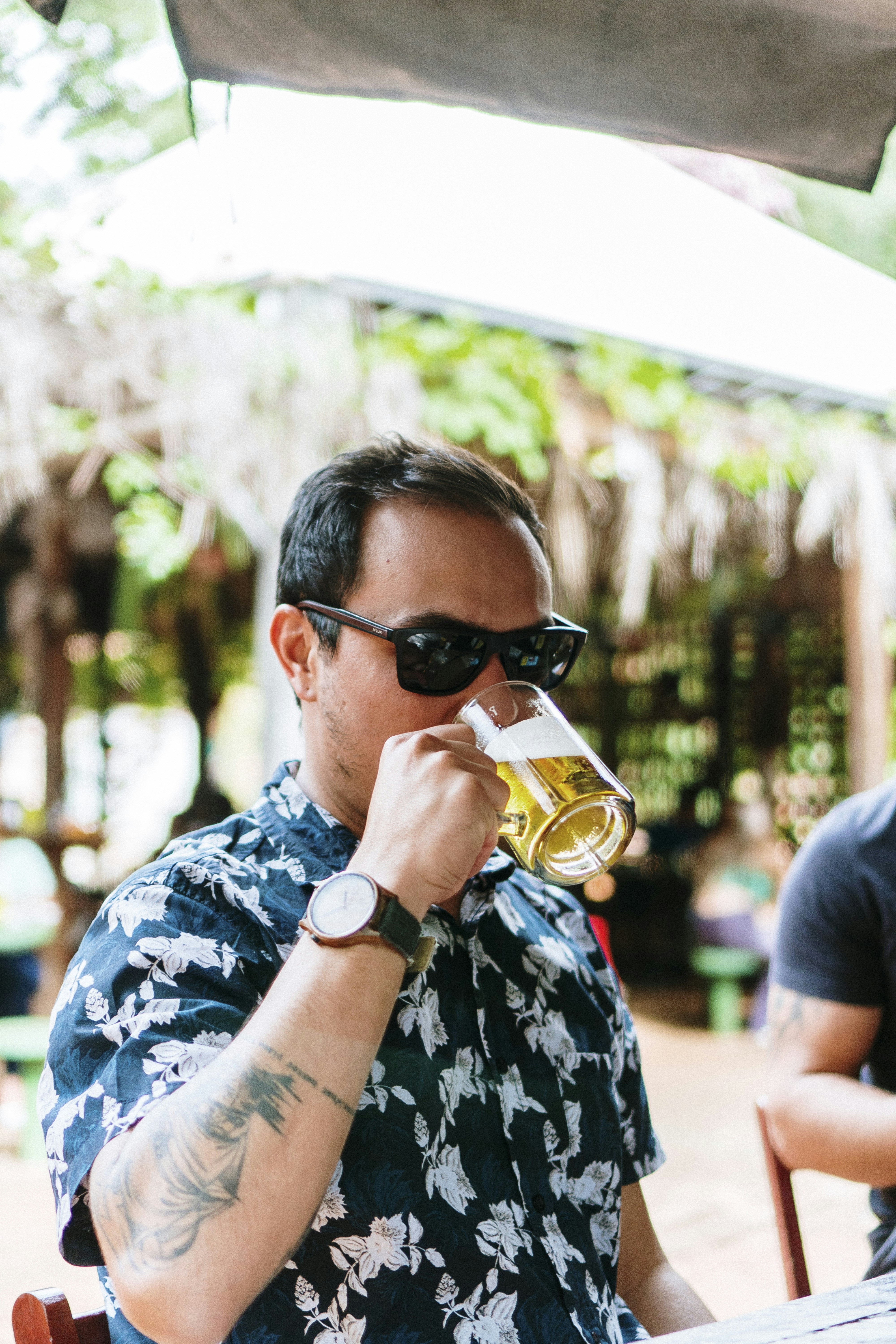 Man in sunglasses enjoying a glass of beer while seated at a lively outdoor setting. Floral shirt adds a vibrant touch to the relaxed atmosphere.