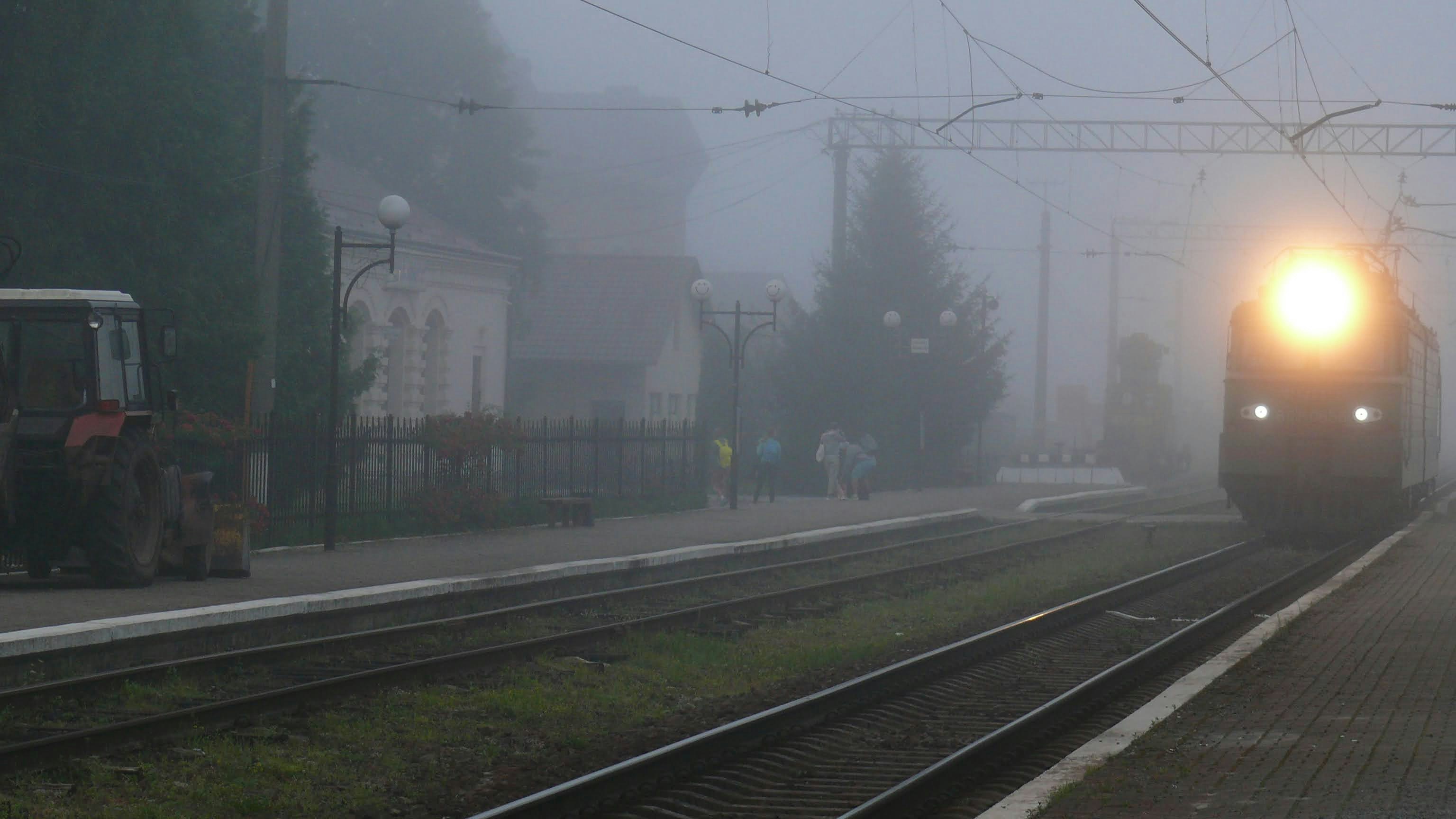 A train emerges from the fog at a quiet station, with a tractor parked nearby and silhouettes of people in the distance. The atmosphere is thick with mist, creating a mysterious ambiance.