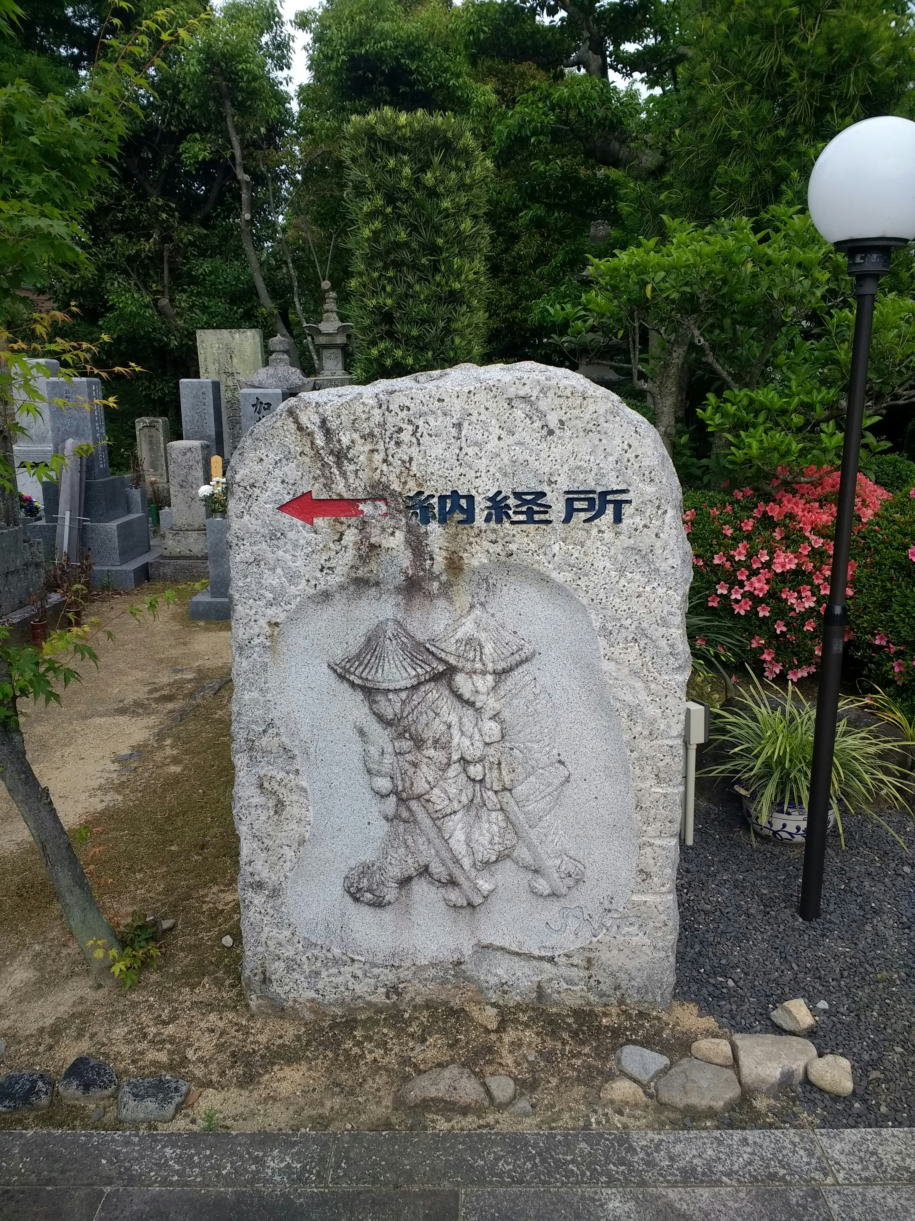 Granite grave marker with relief carving of two figures, blue Japanese text and a red arrow. Set in a tidy garden cemetery with a lamp post and flowering shrubs.