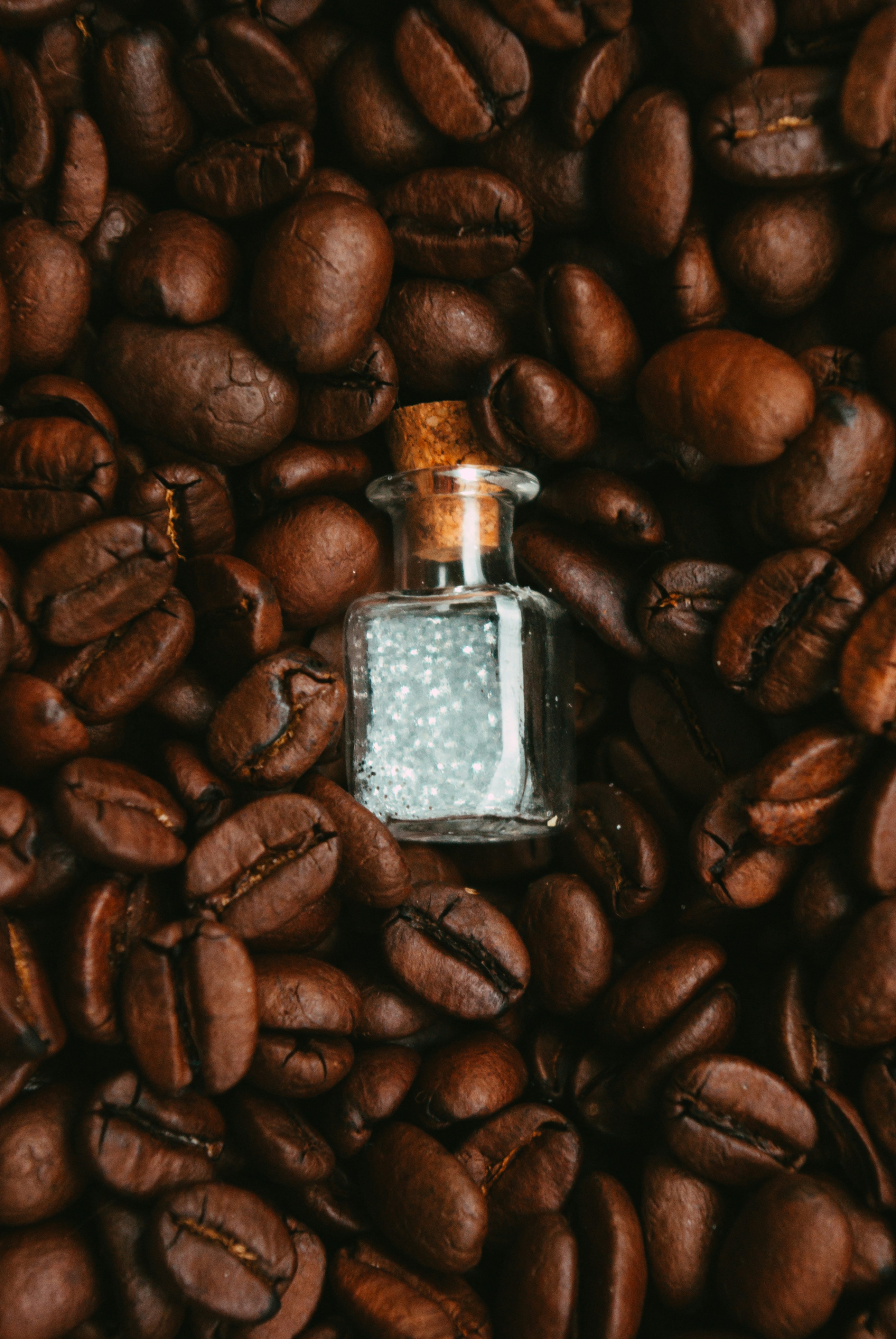 Close-up photograph of a small corked glass bottle nestled among roasted coffee beans, with crystalline contents visible inside.