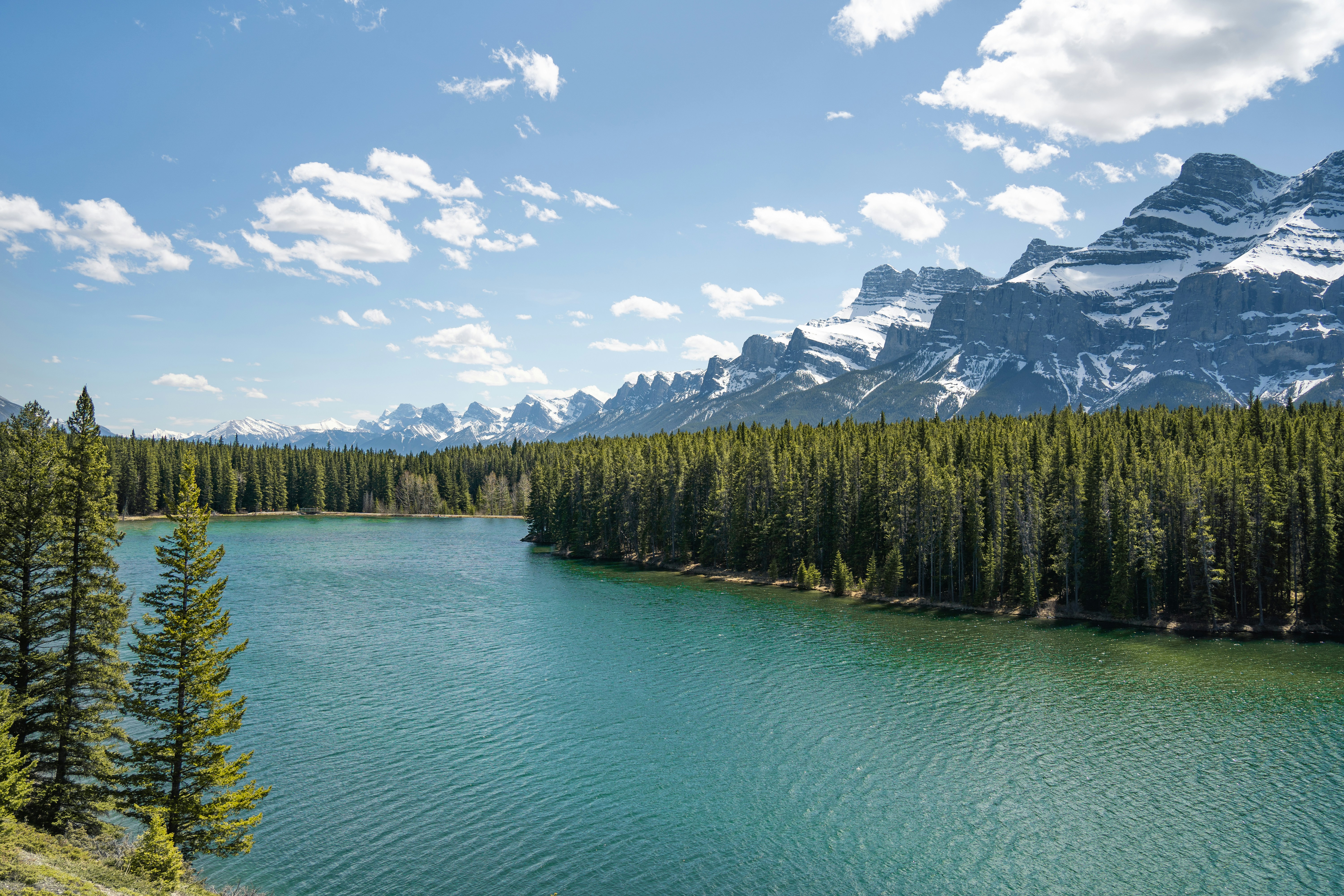 Turquoise lake surrounded by dense evergreen forest with snow-capped mountains under a clear blue sky.