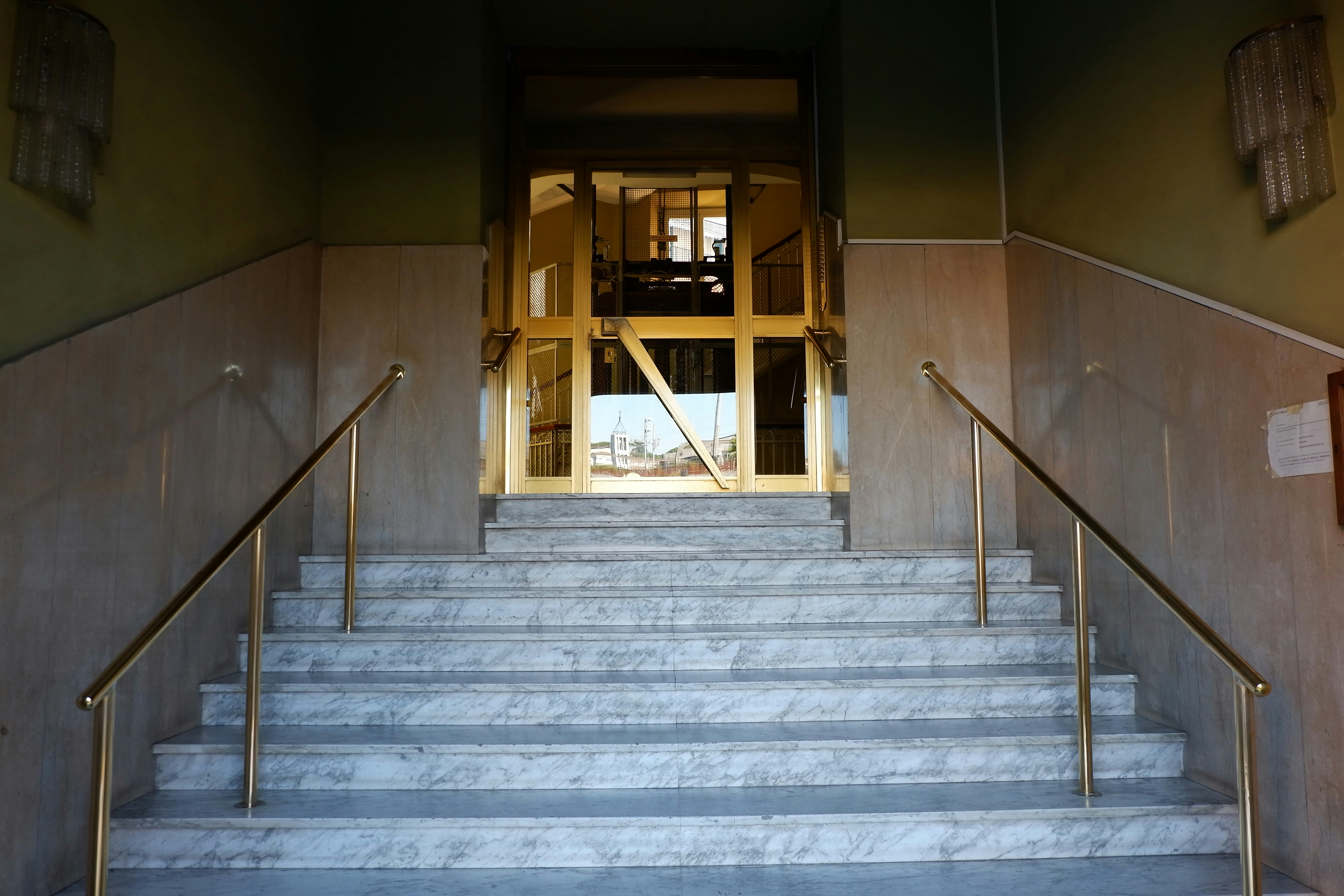 A photograph of a marble staircase ascending to a brightly lit doorway framed by glass panels; brass handrails lead the eye toward the lobby beyond.