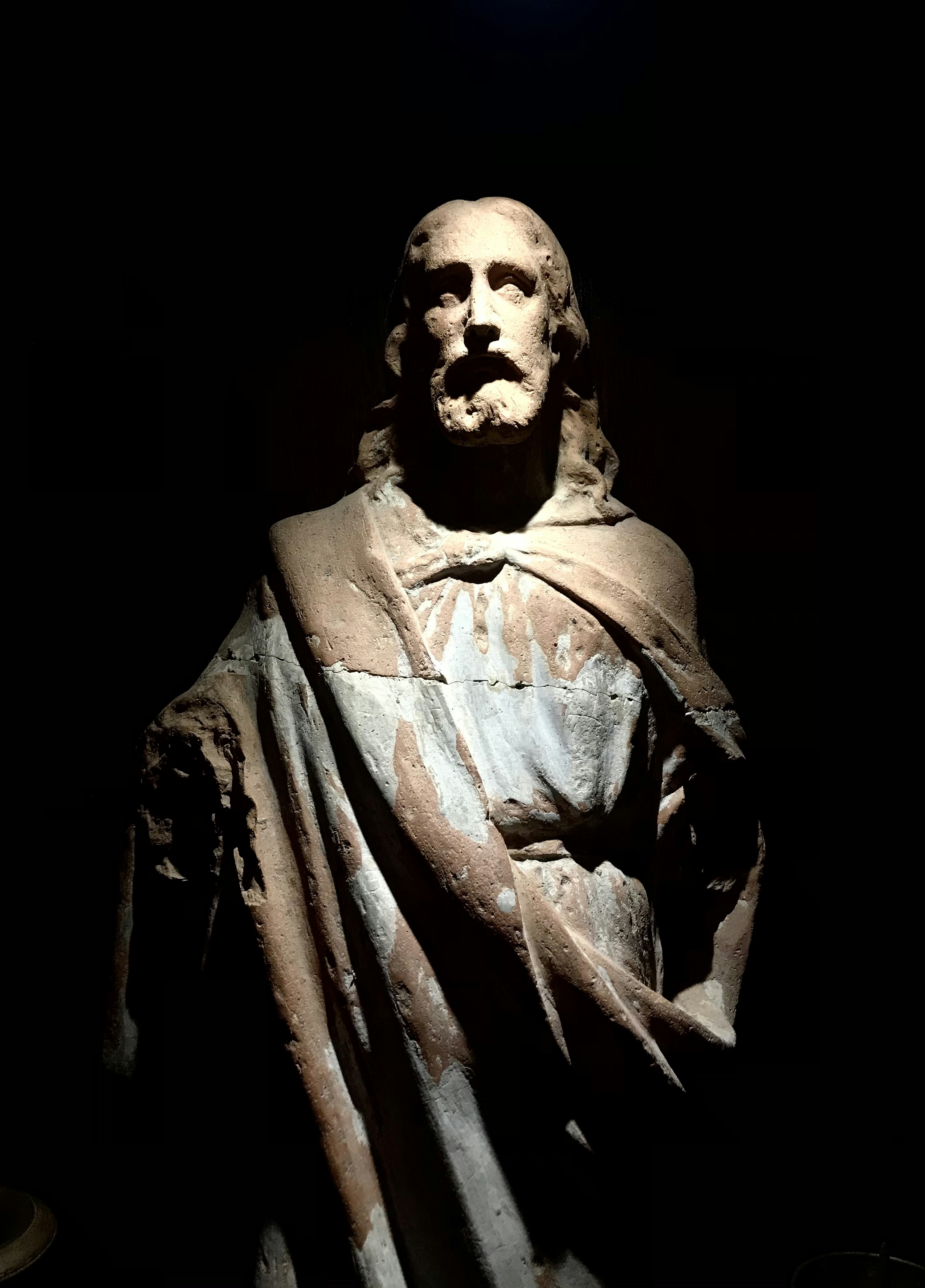 A weathered statue draped in a cloak, illuminated dramatically against a dark backdrop, revealing intricate details and textures.