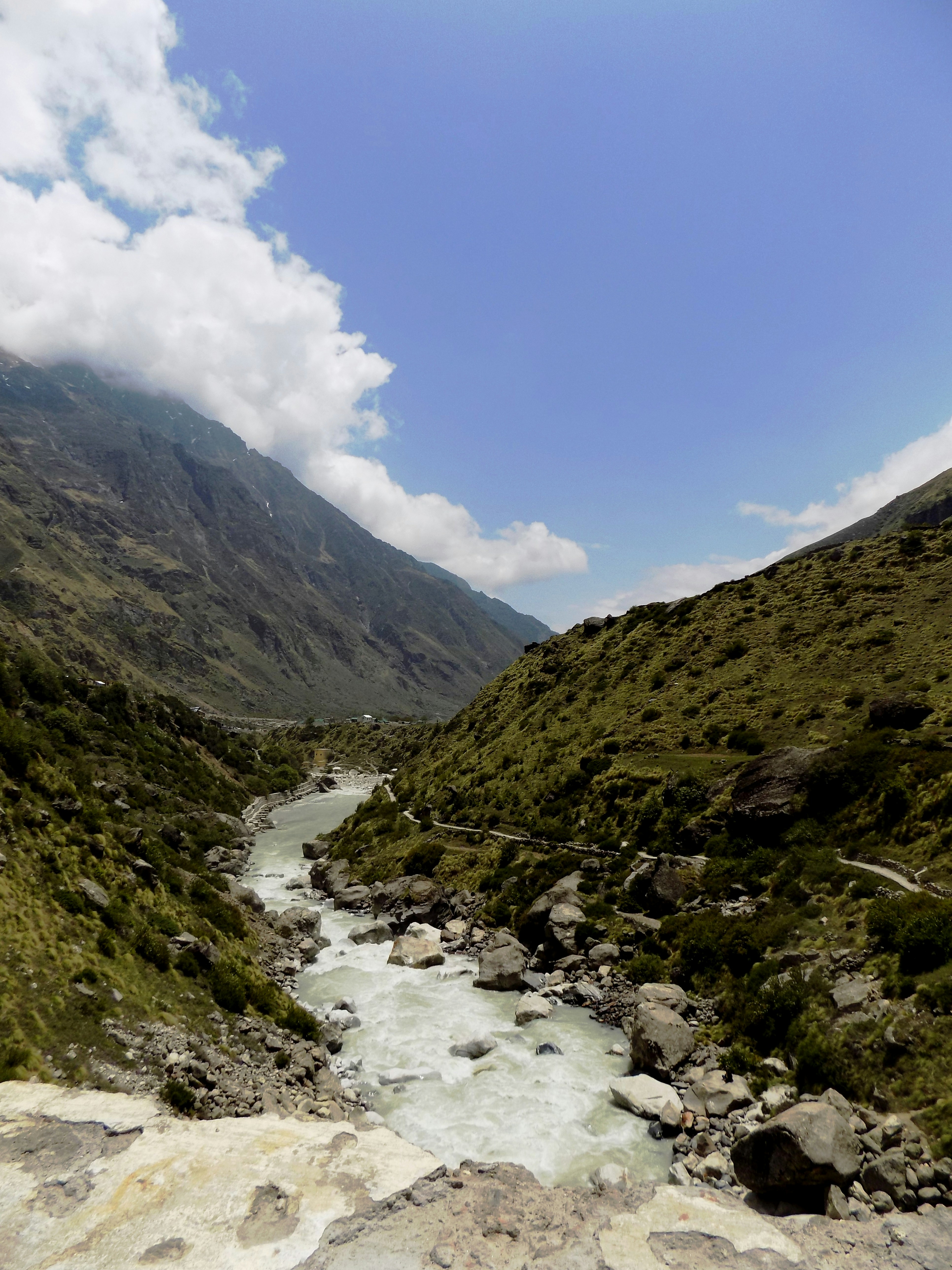 Landscape photograph of a rugged mountain valley with a fast-flowing river cutting through rocky banks. A blue sky and scattered clouds crown the scene.