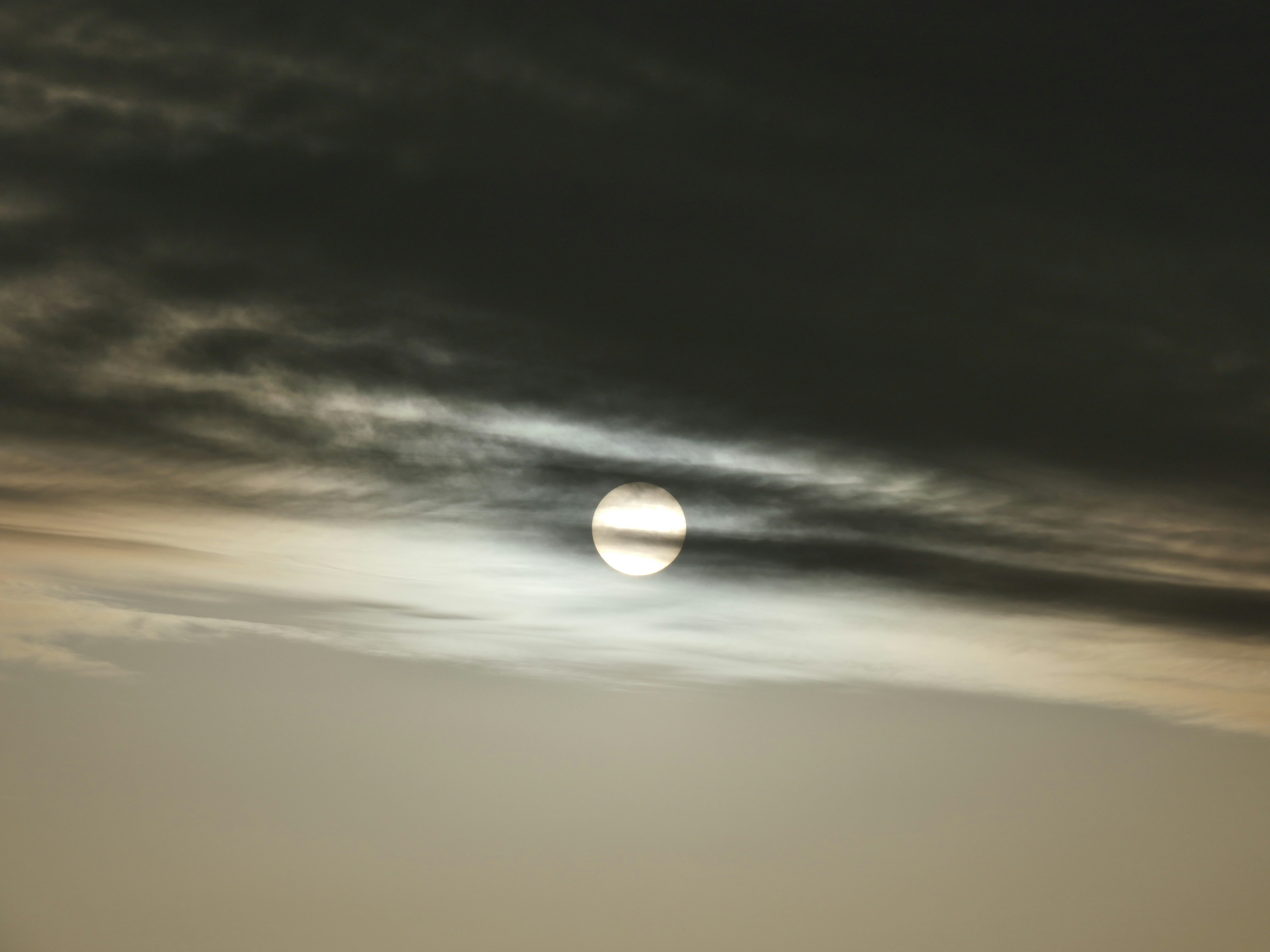 Sun partially obscured by clouds, casting a soft glow in a muted sky. The interplay of light and shadow creates a serene atmosphere.