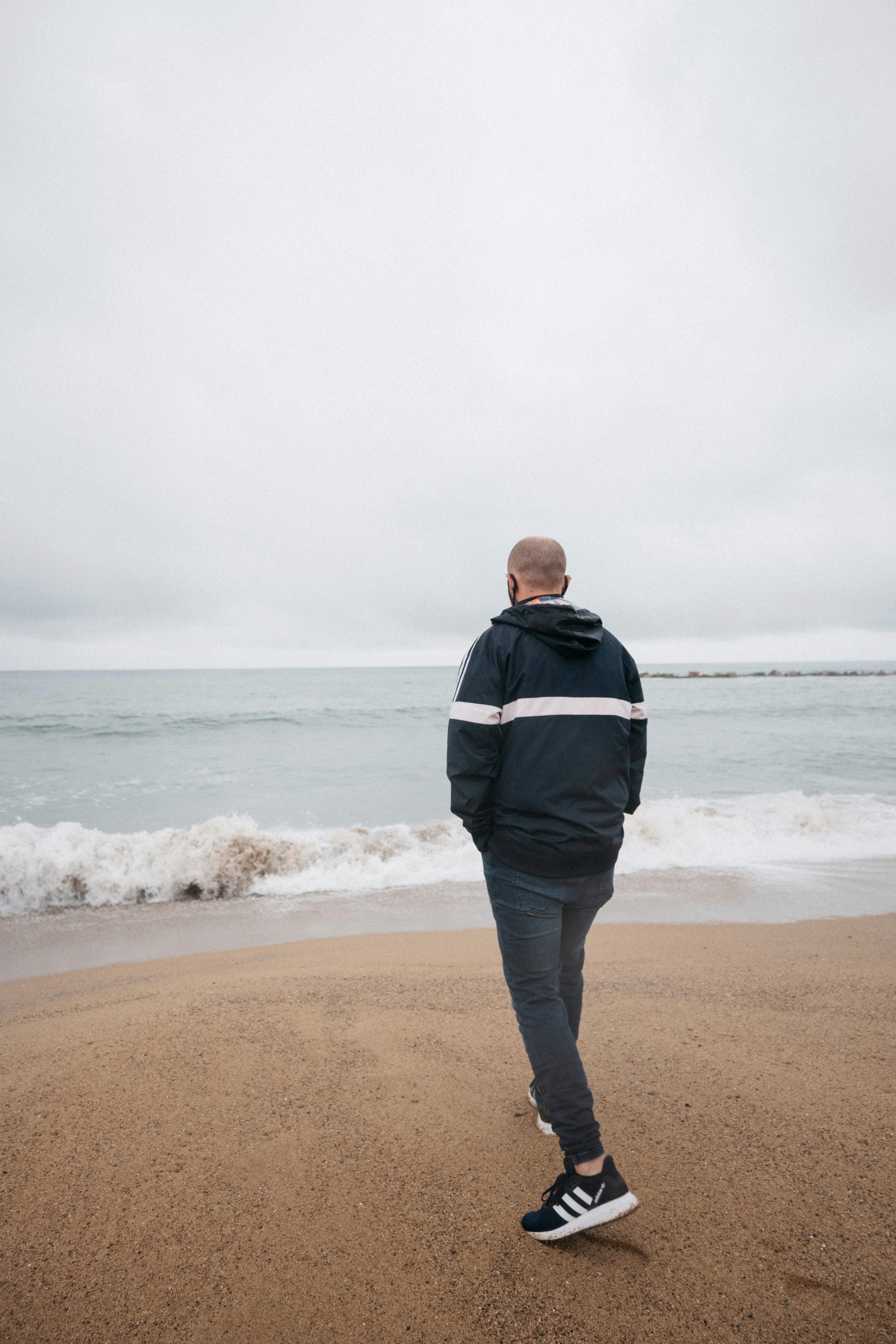 A person in a dark jacket walks along a sandy beach, gazing out at the calm sea under a cloudy sky.