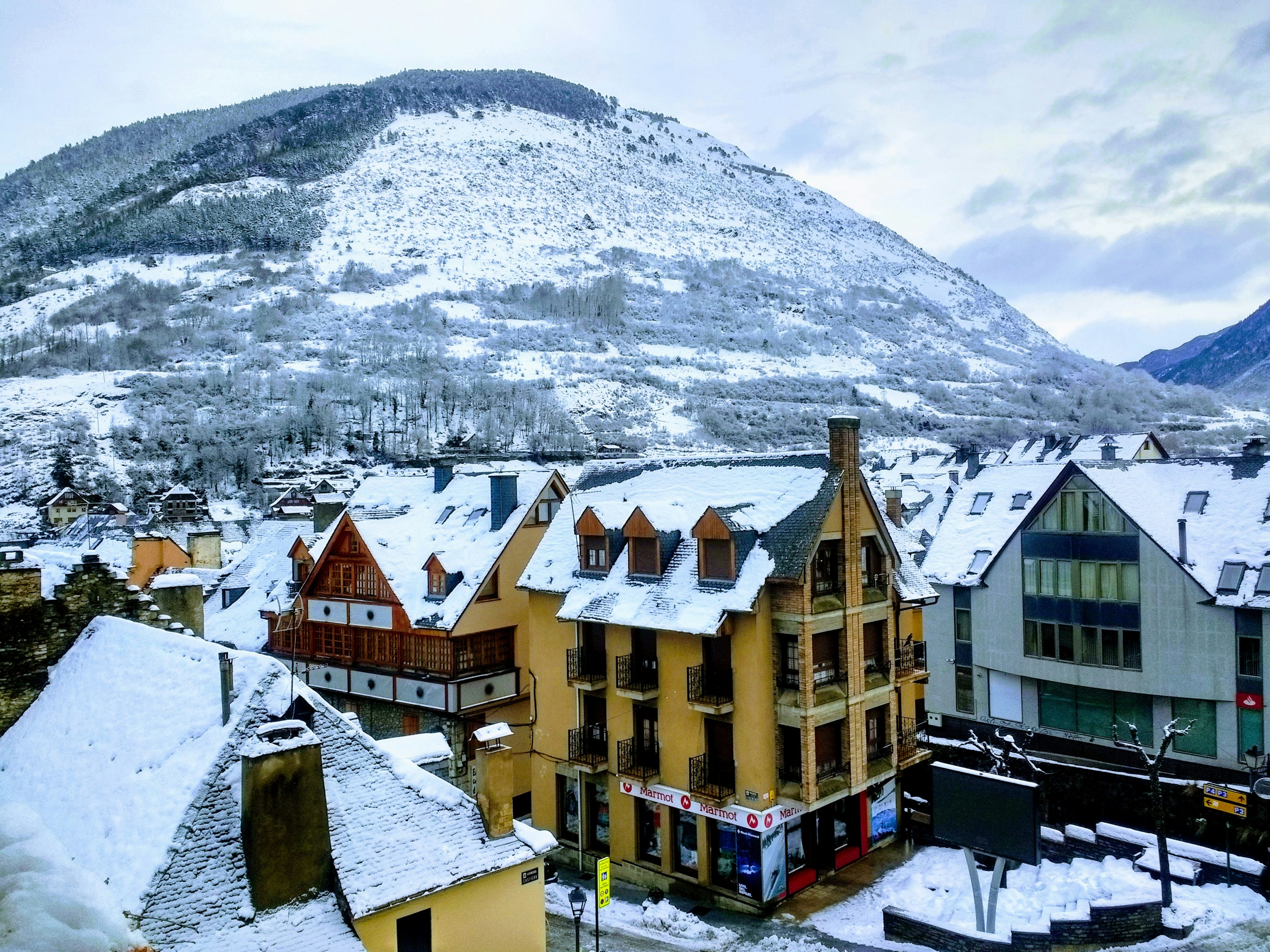 Snow-covered rooftops in a quaint village nestled against a vast mountain landscape under a cloudy sky.