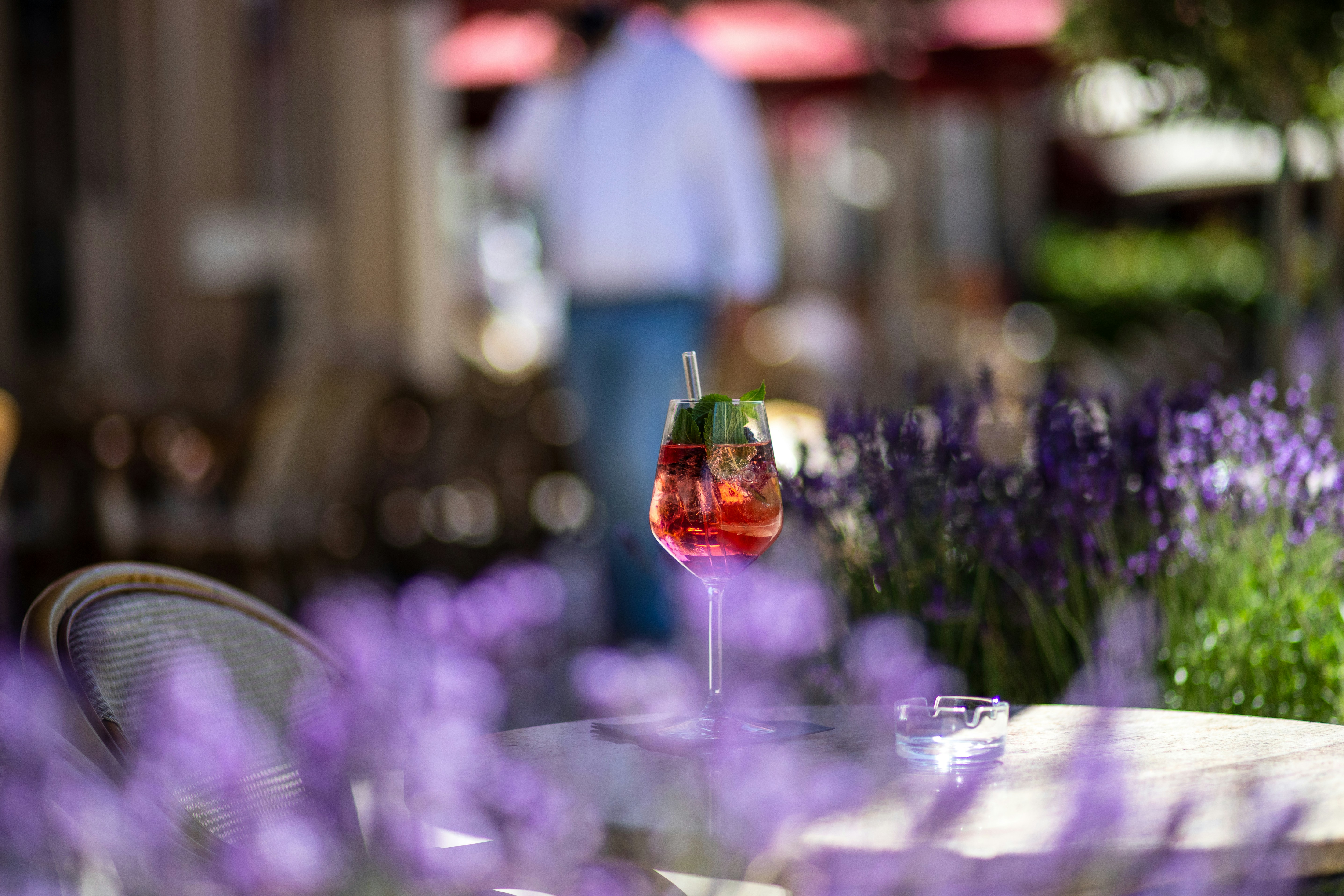 Wine glass filled with red liquid on a sunlit table amidst purple lavender blossoms.