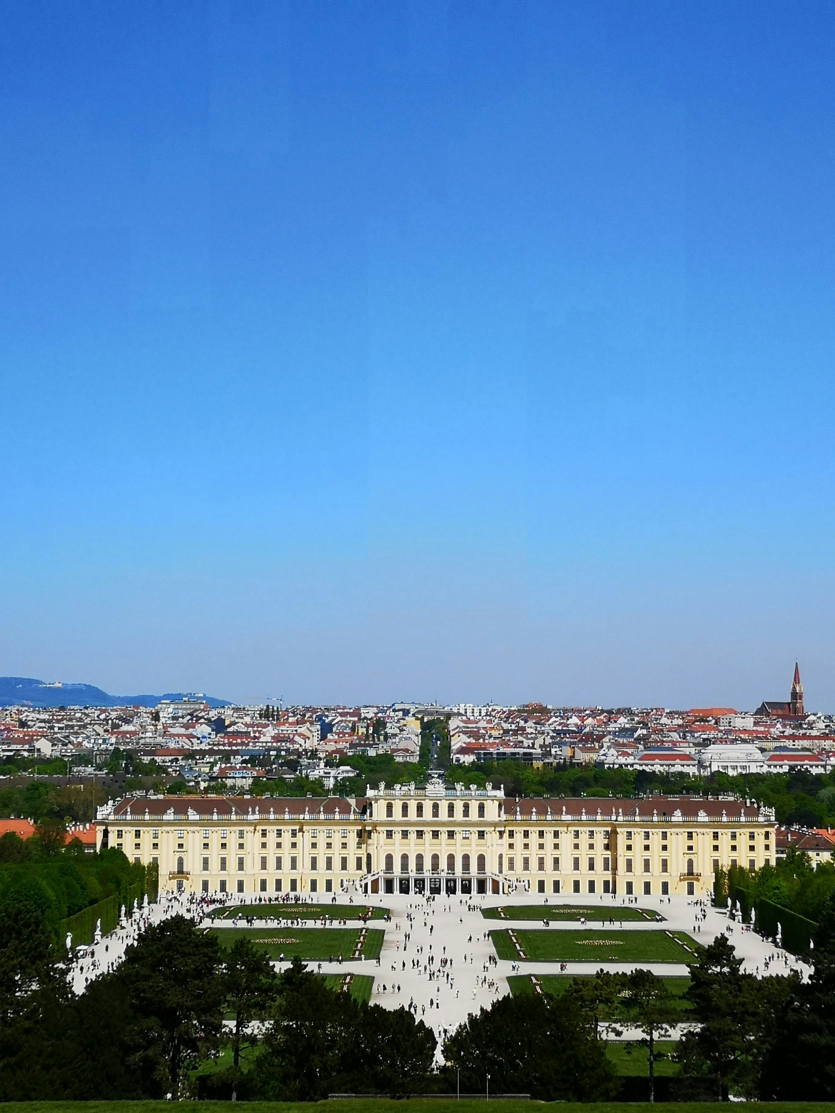 Schönbrunn Palace stands majestically amidst lush gardens and urban landscape under a clear blue sky.