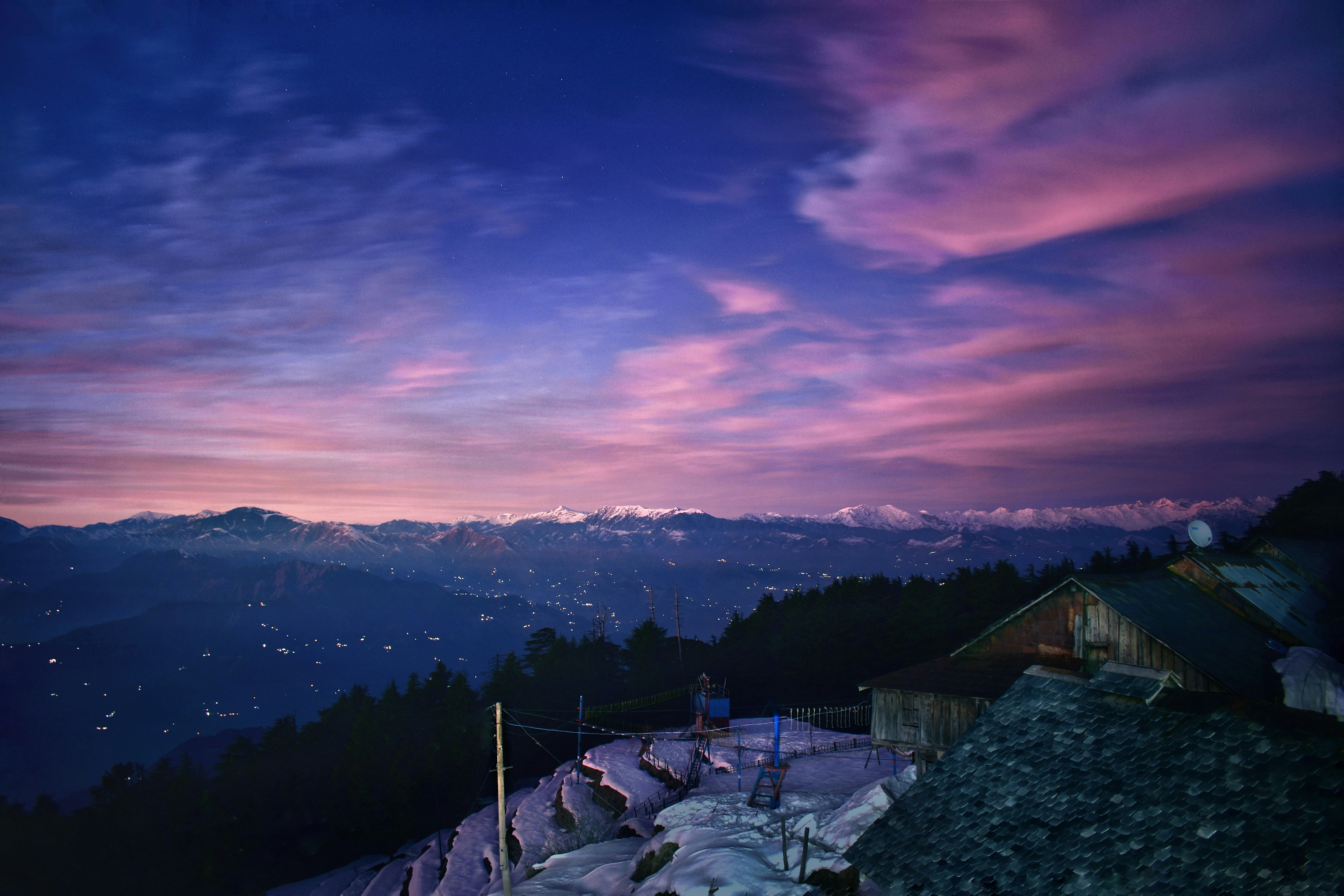 Colorful dawn sky over snow-dusted mountains with a rustic rooftop in the foreground.
