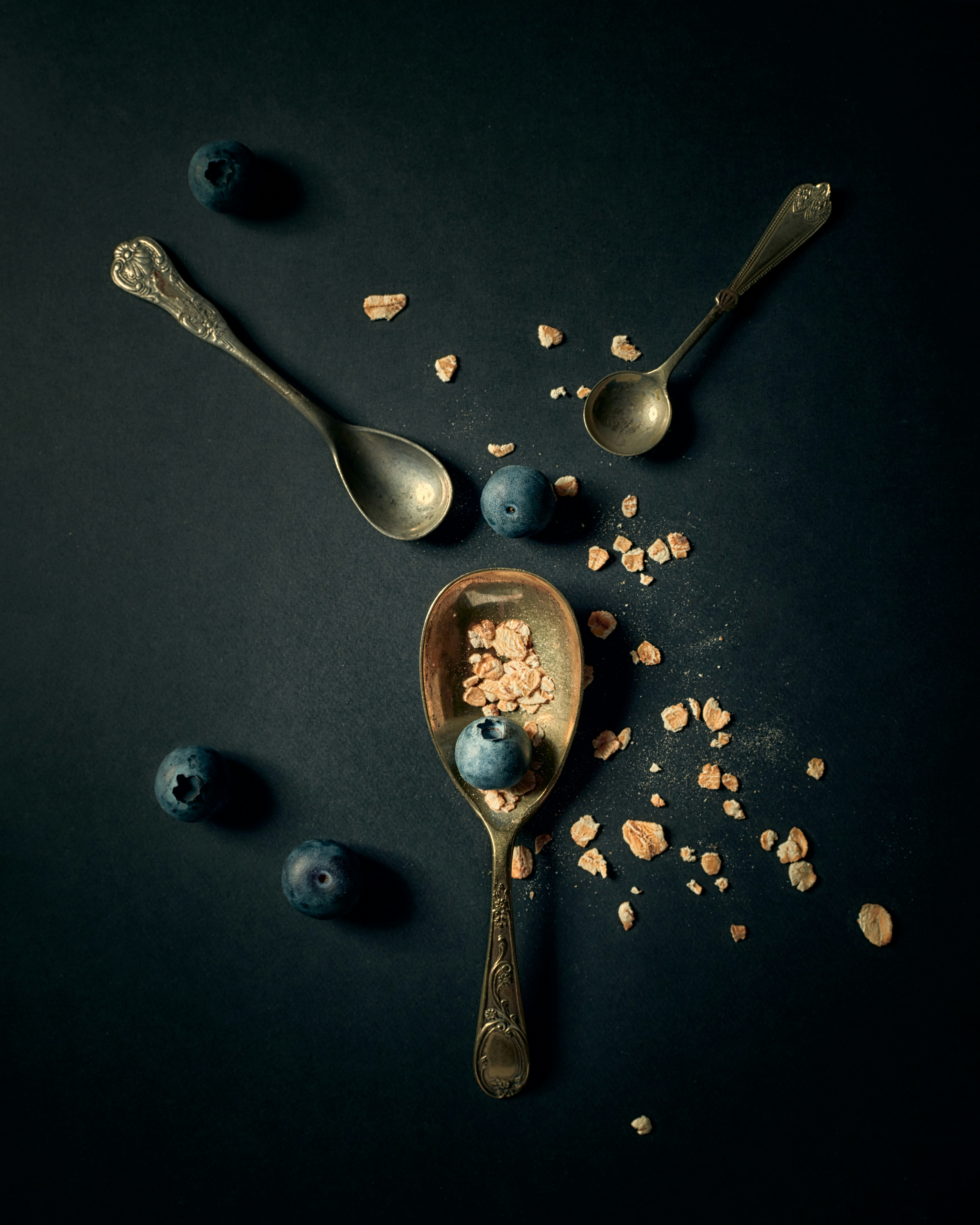 A creative arrangement of vintage silver spoons and blueberries scattered over a dark surface, with granola hints adding texture.