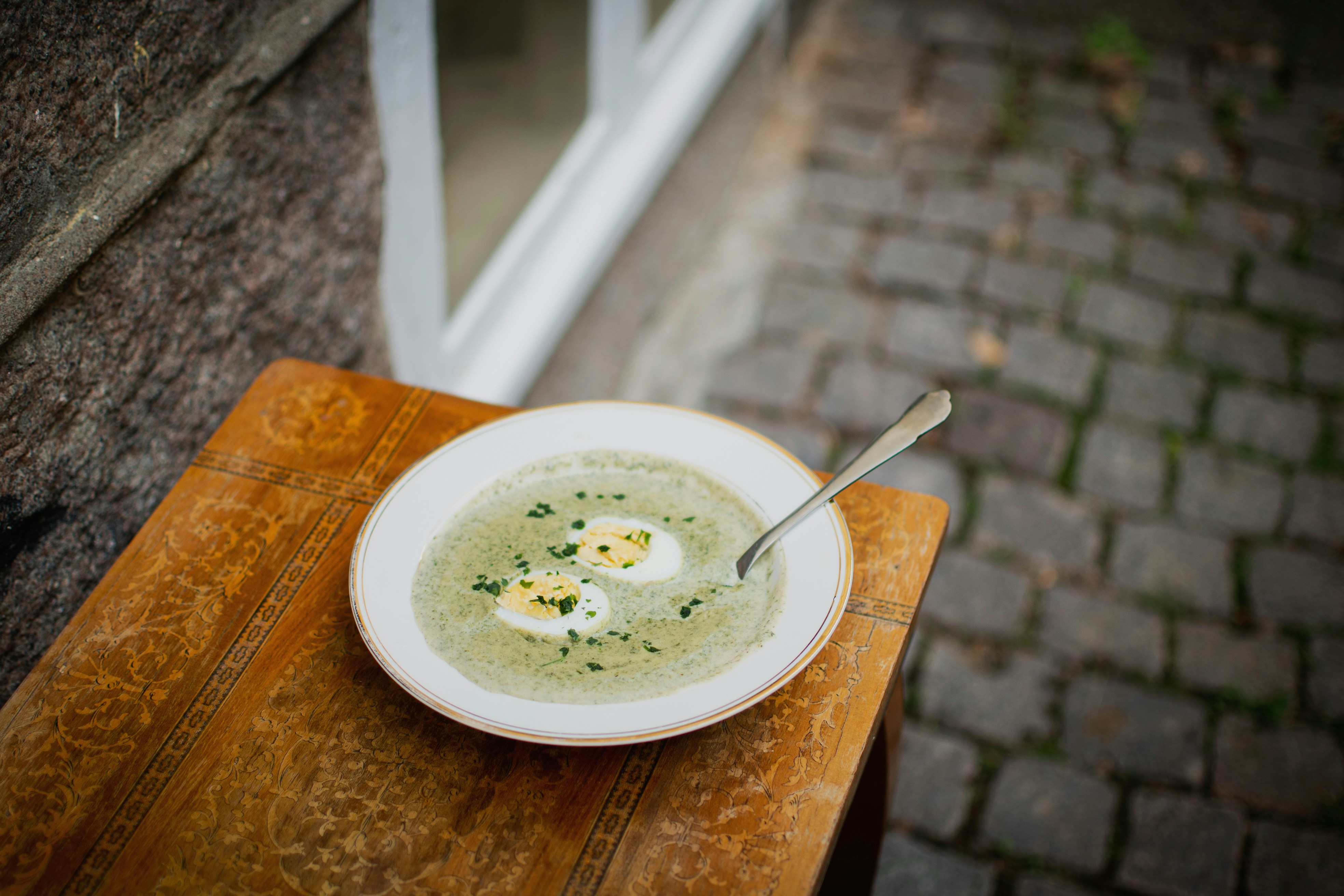 Creamy green soup garnished with soft-boiled eggs and herbs, elegantly presented on a wooden table beside cobblestone pavement.