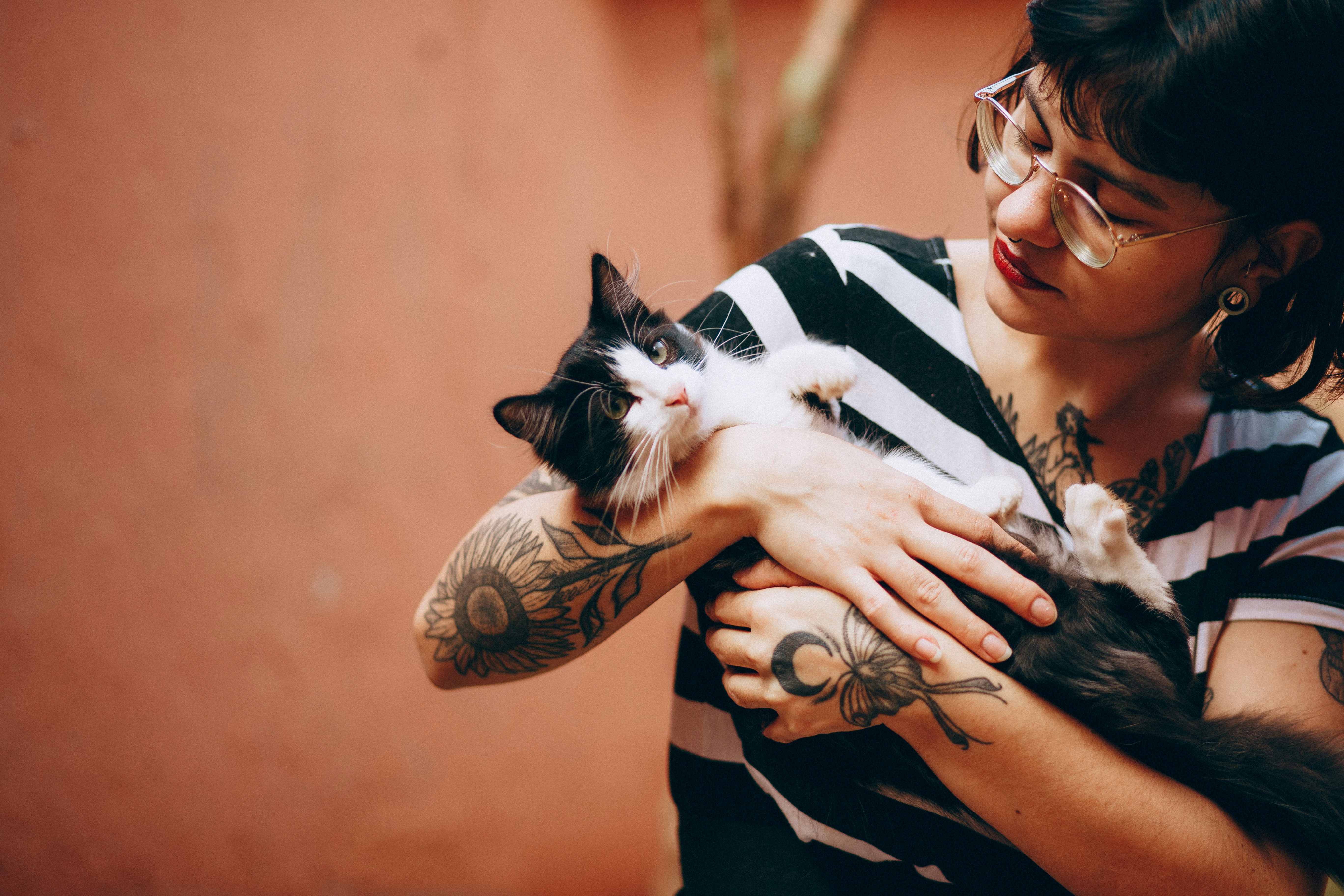 A woman with tattoos and glasses cradles a black-and-white cat against a warm, peach-toned background, conveying tenderness in a candid portrait.