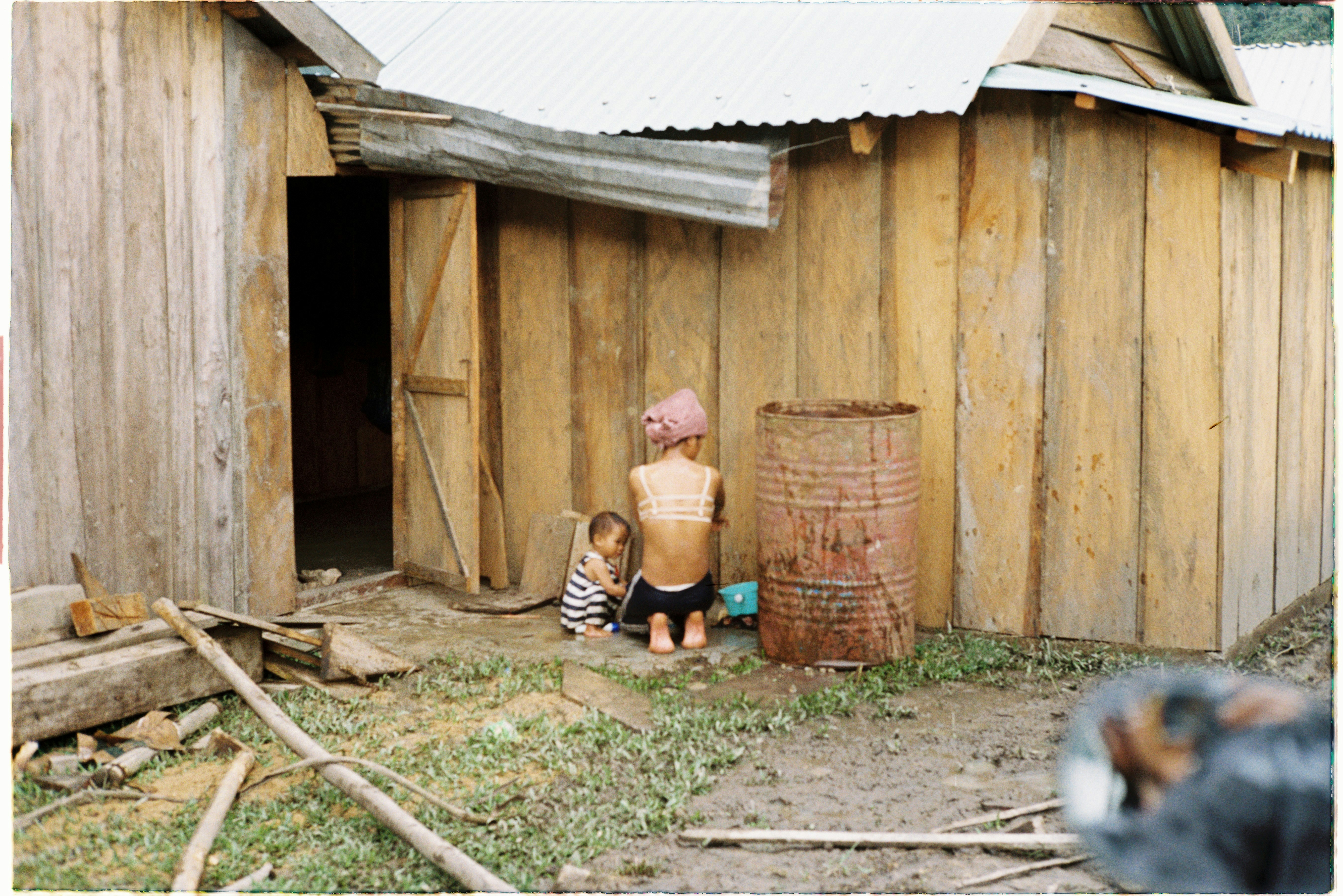 Children near a weathered wooden hut with a rusty barrel
