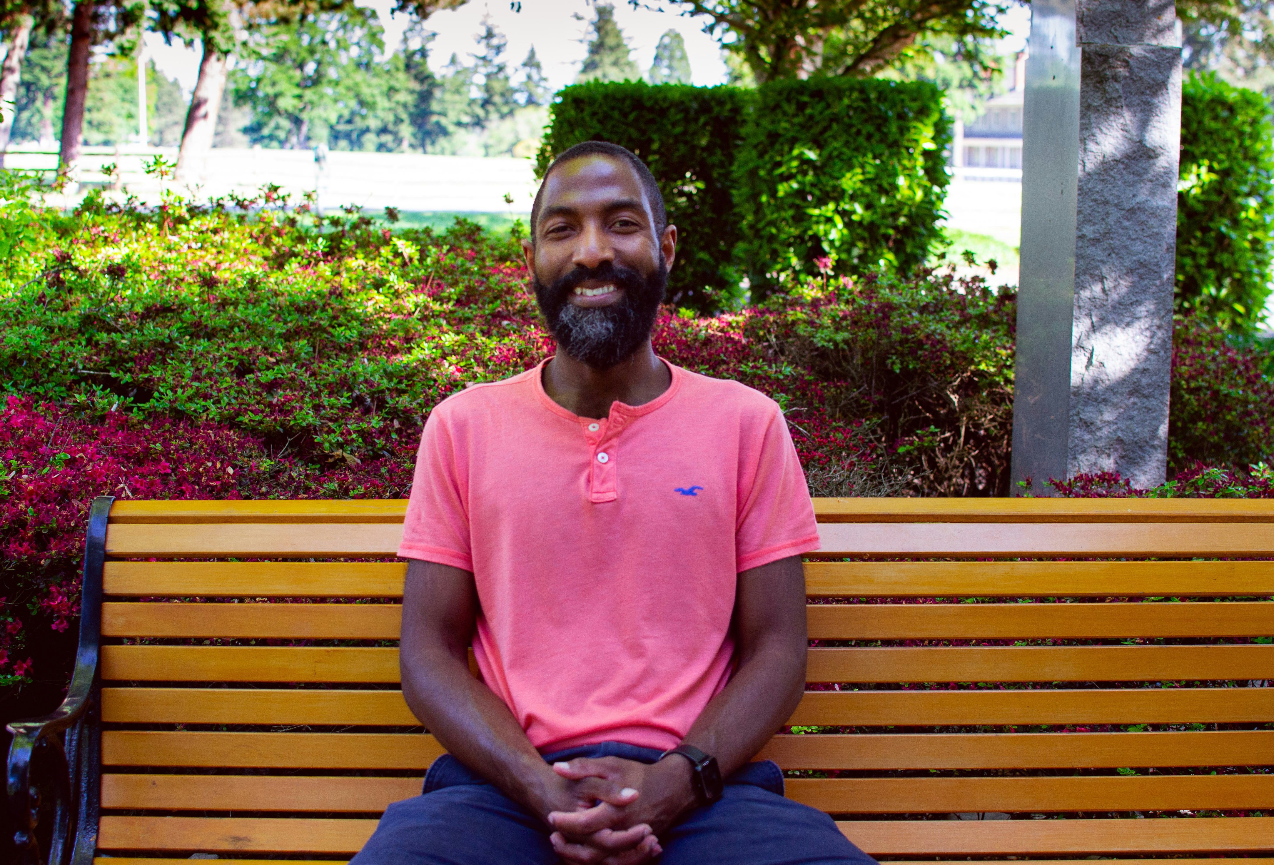 Smiling man seated on a wooden bench surrounded by lush greenery and colorful flowers in a park.