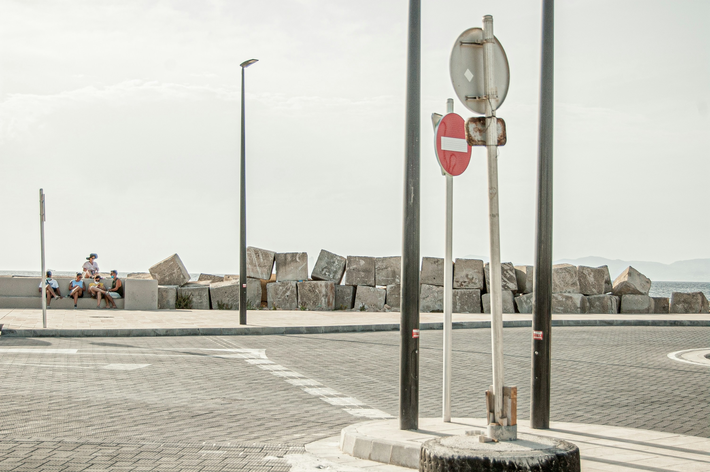 Two individuals relax on a coastal promenade, framed by traffic signs and a backdrop of large stone blocks. The scene conveys a sense of calm in an urban setting.