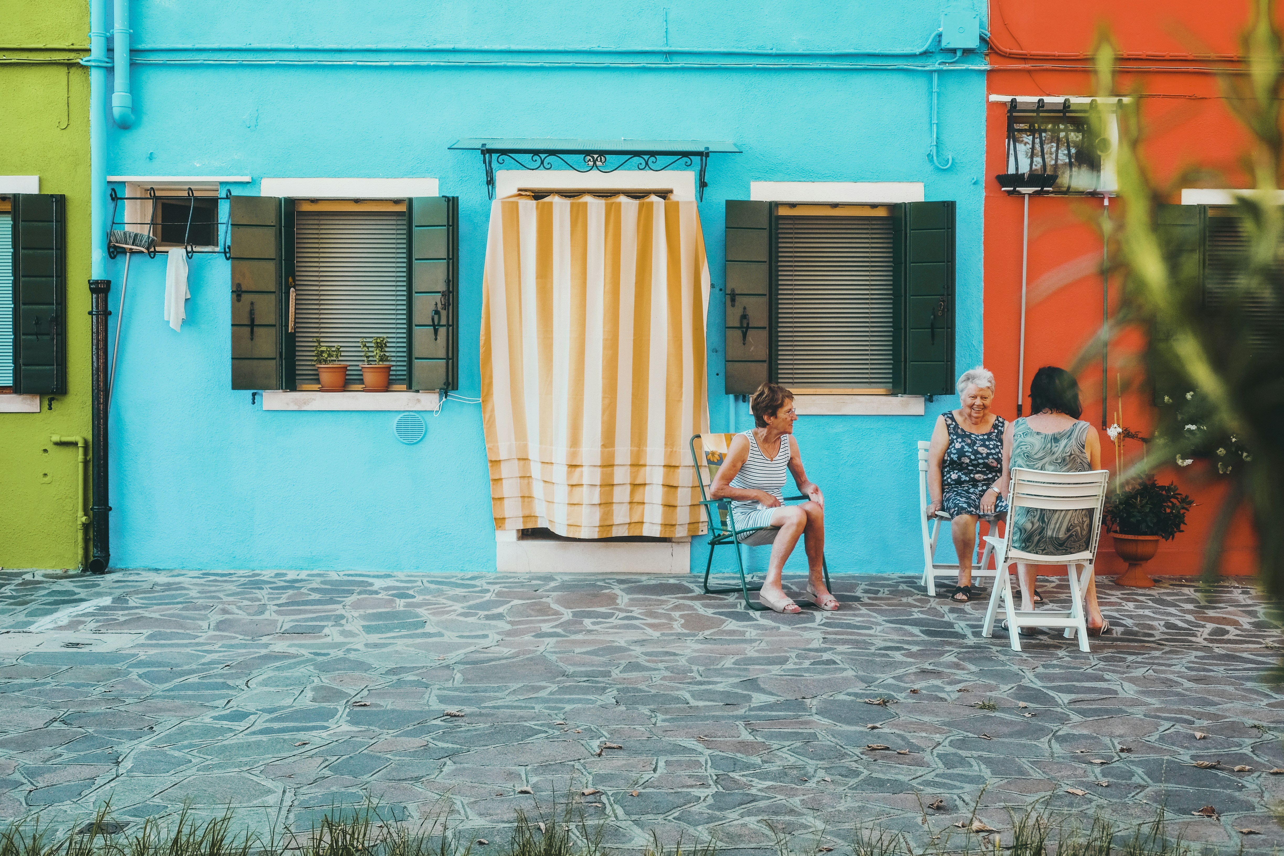 Three women engaged in conversation outside a vibrant blue and orange building, with a striped curtain adding a playful element.
