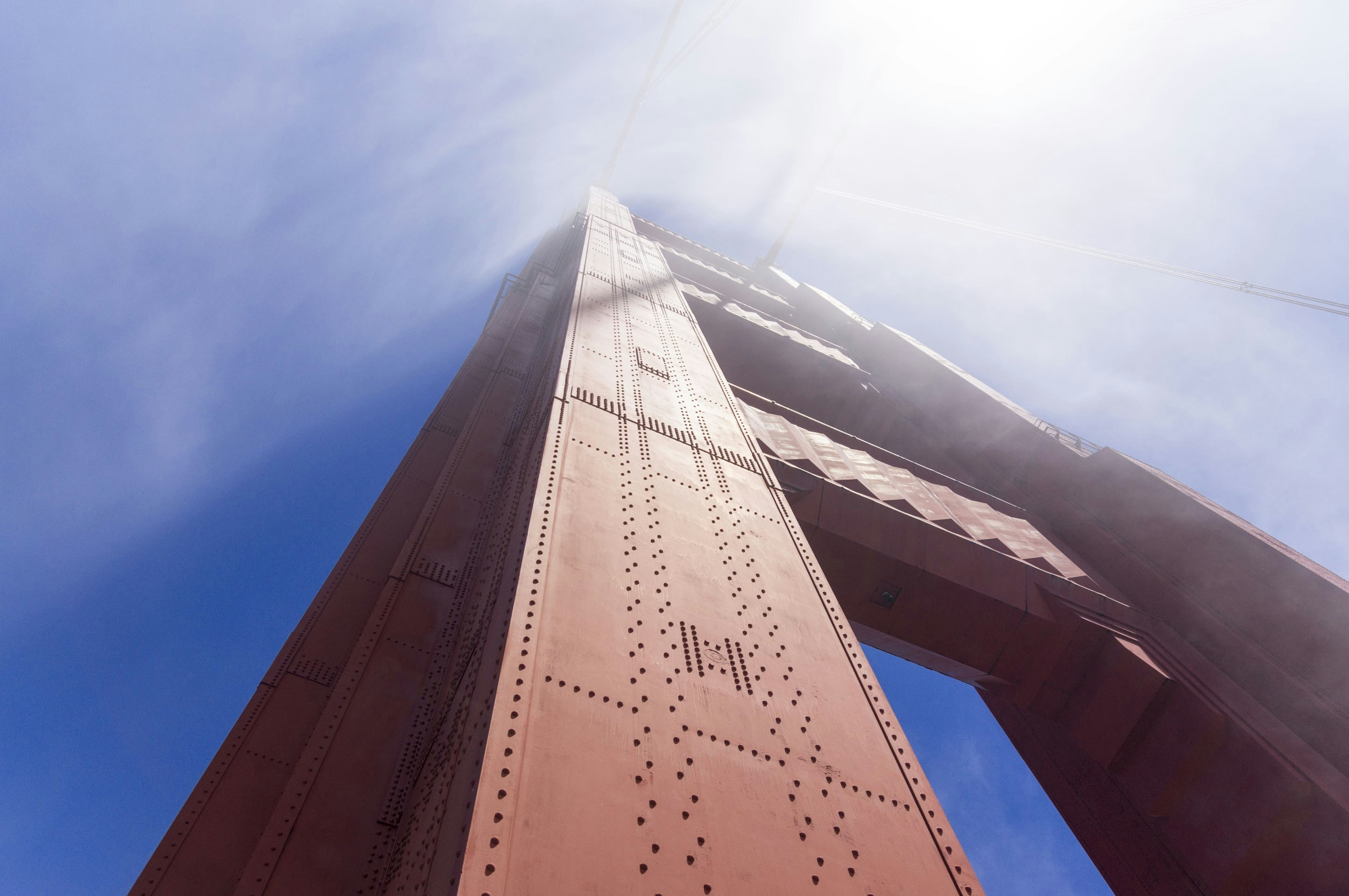 A low-angle view of a rust-colored steel arch tower stretching toward a bright sky; sun glare adds a halo around the top, emphasizing the structure’s riveted surface and geometric lines. This is a photograph of a bridge girder, highlighting industrial architecture.