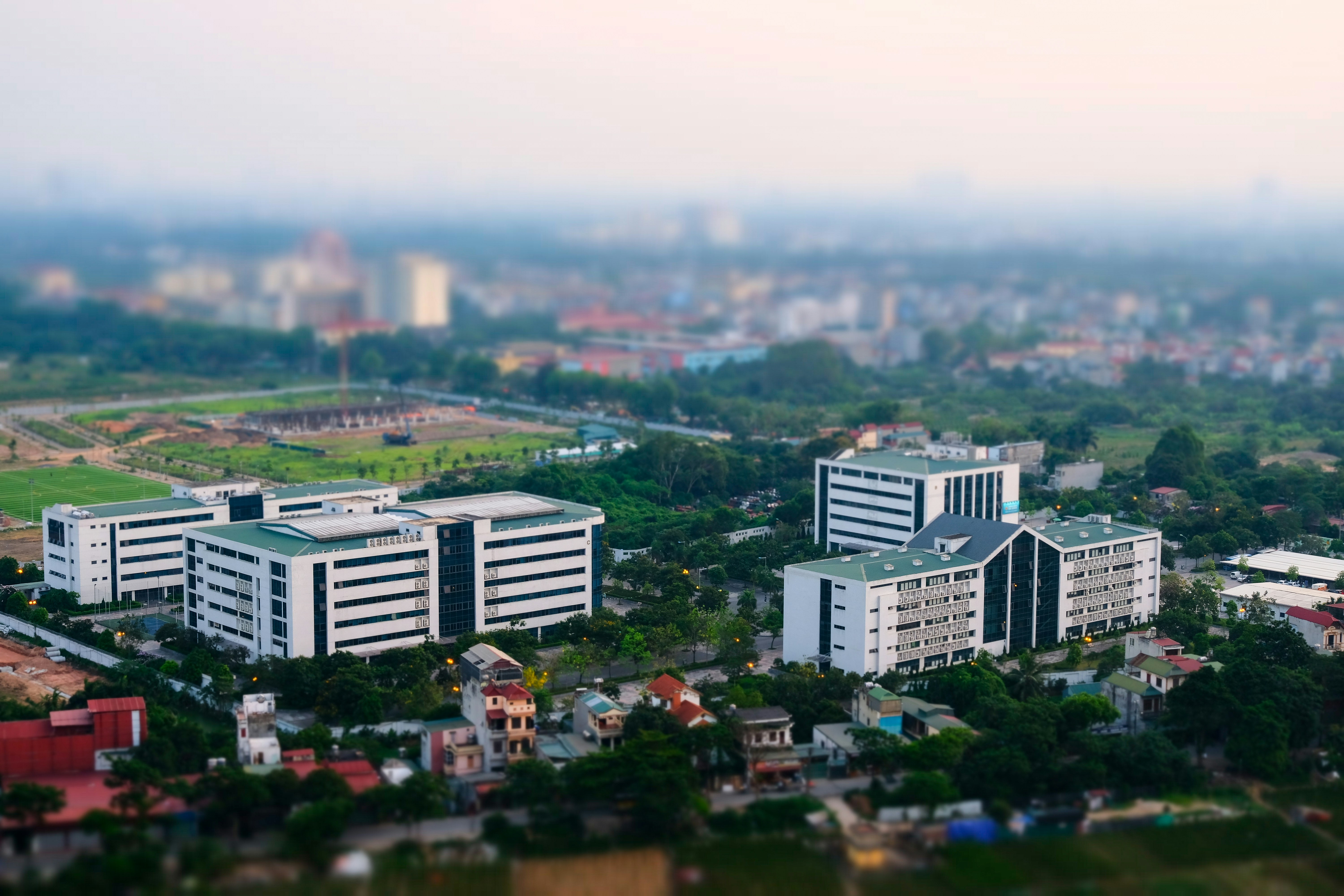 Tilt-shift view of a modern campus surrounded by lush greenery and distant city skyline.