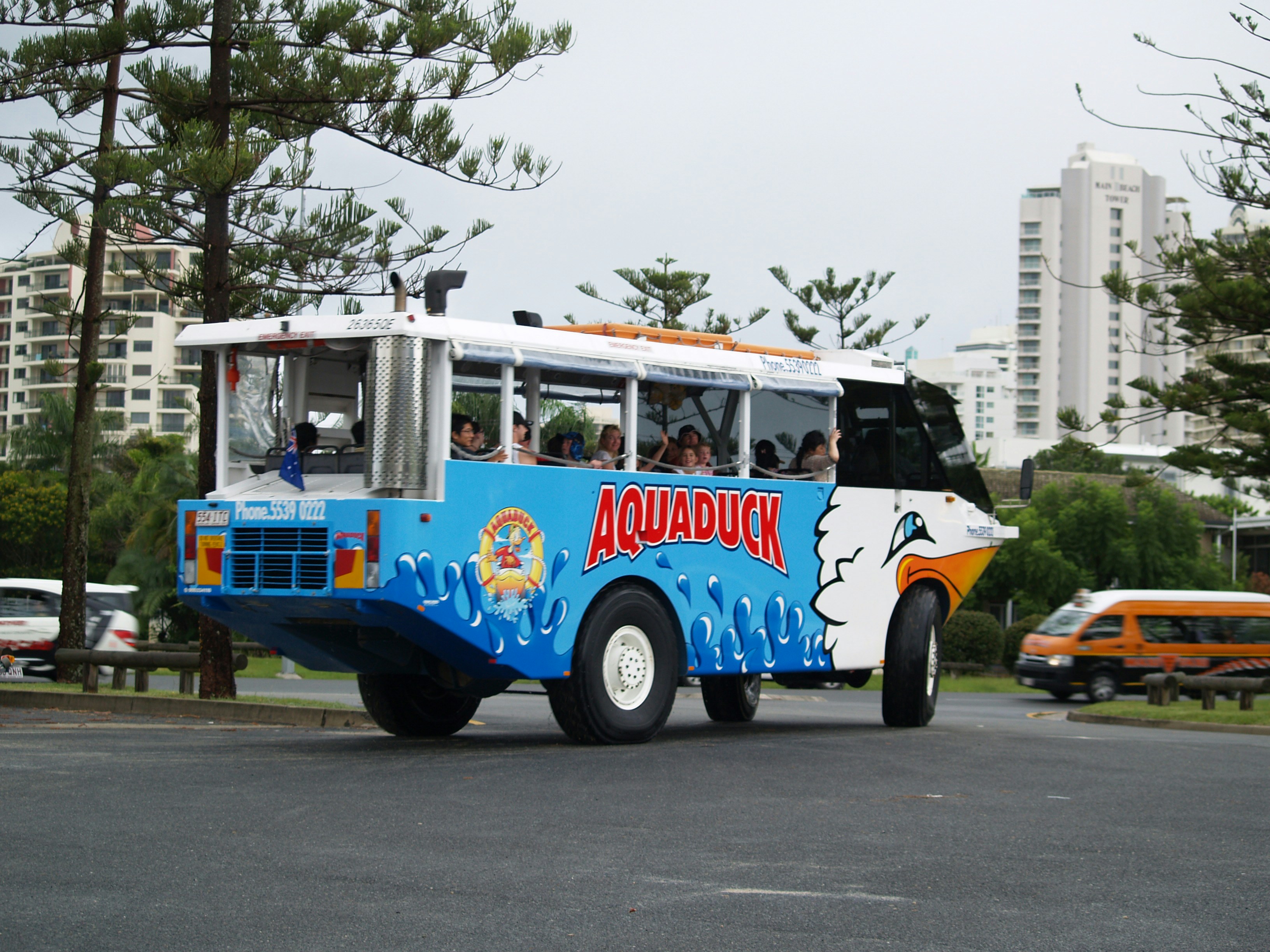 Colorful Aquaduck amphibious vehicle parked in a scenic area, with passengers enjoying the ride. The backdrop features urban buildings and greenery.