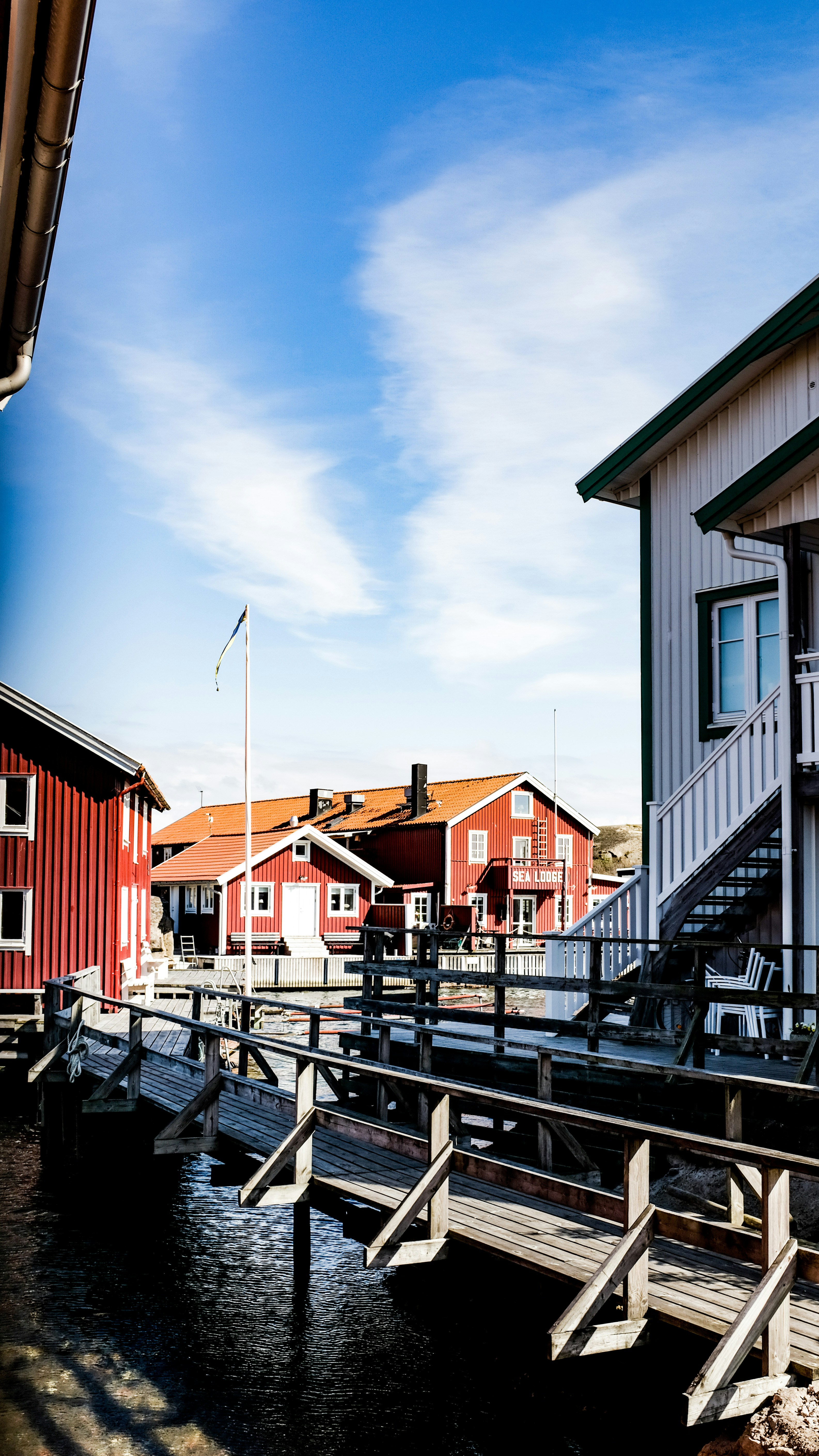 Charming wooden buildings in vibrant red and white line a scenic harbor, with a wooden walkway leading through the tranquil waters.