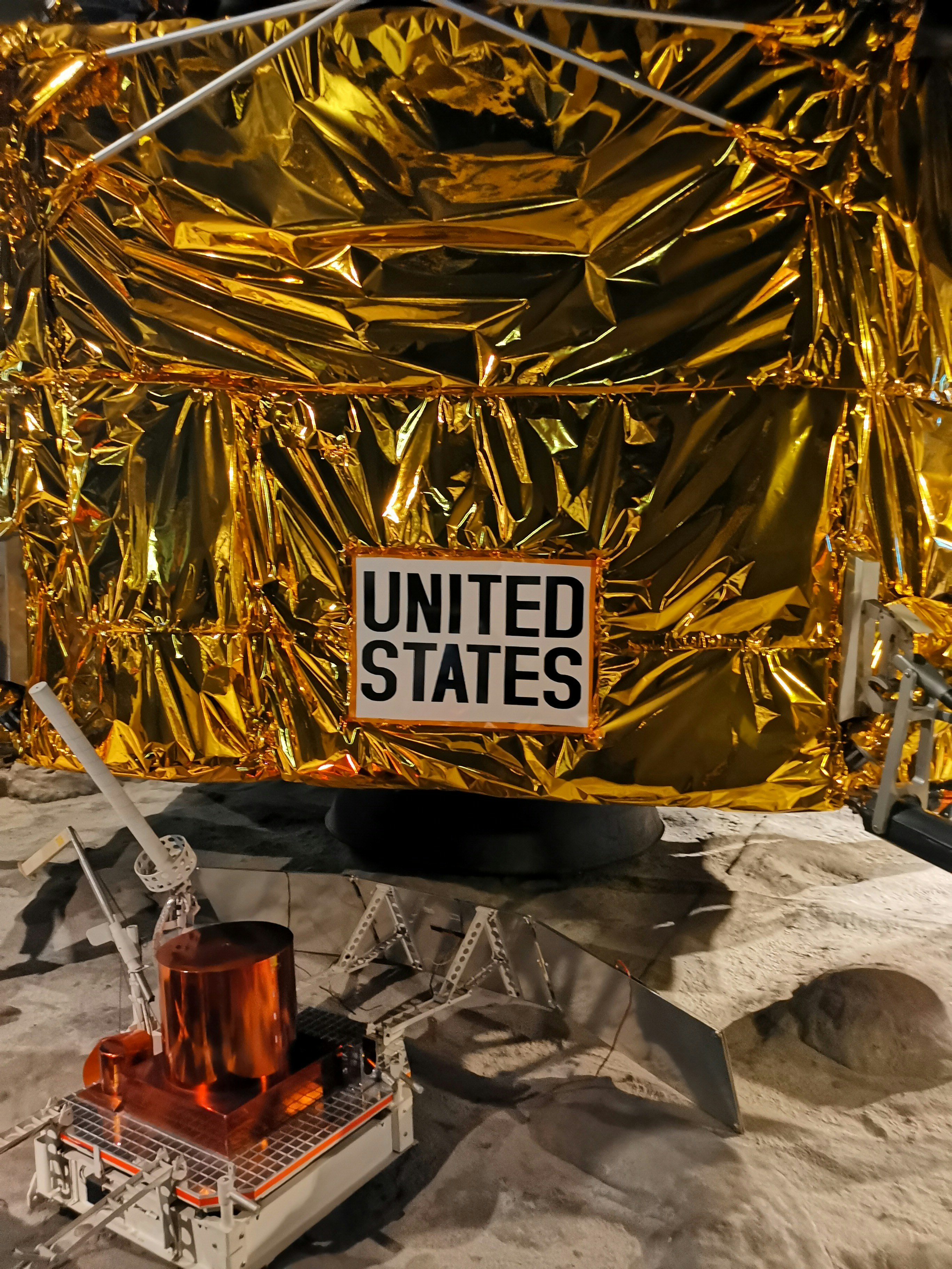 Gold-foil lunar lander model sits on a dusty surface with a United States sign, forming a staged lunar diorama.