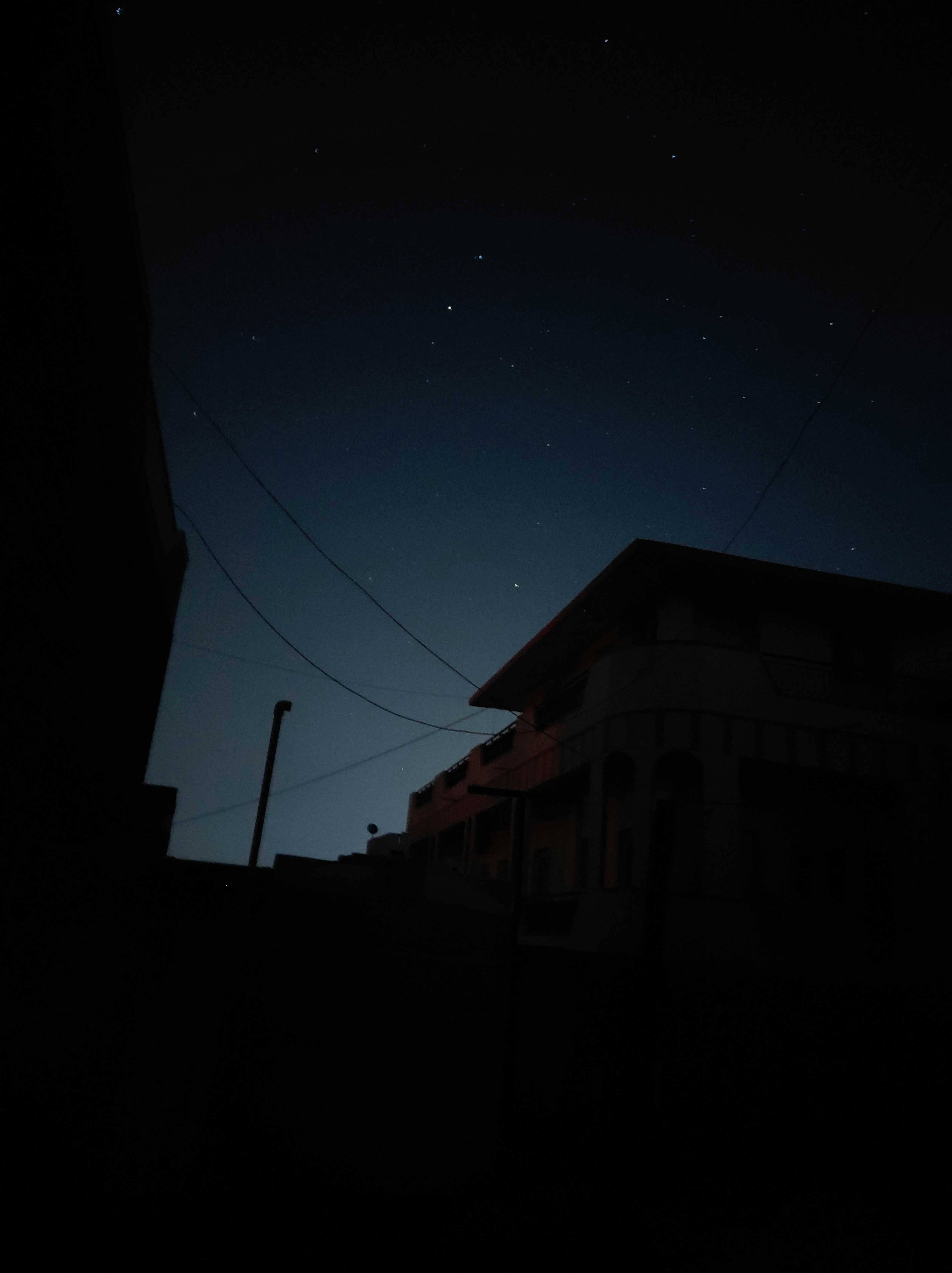 Nighttime urban silhouette beneath a star-filled sky, with power lines crossing the frame and low buildings in shadow. The image captures a calm, blue-tinged nocturnal cityscape.