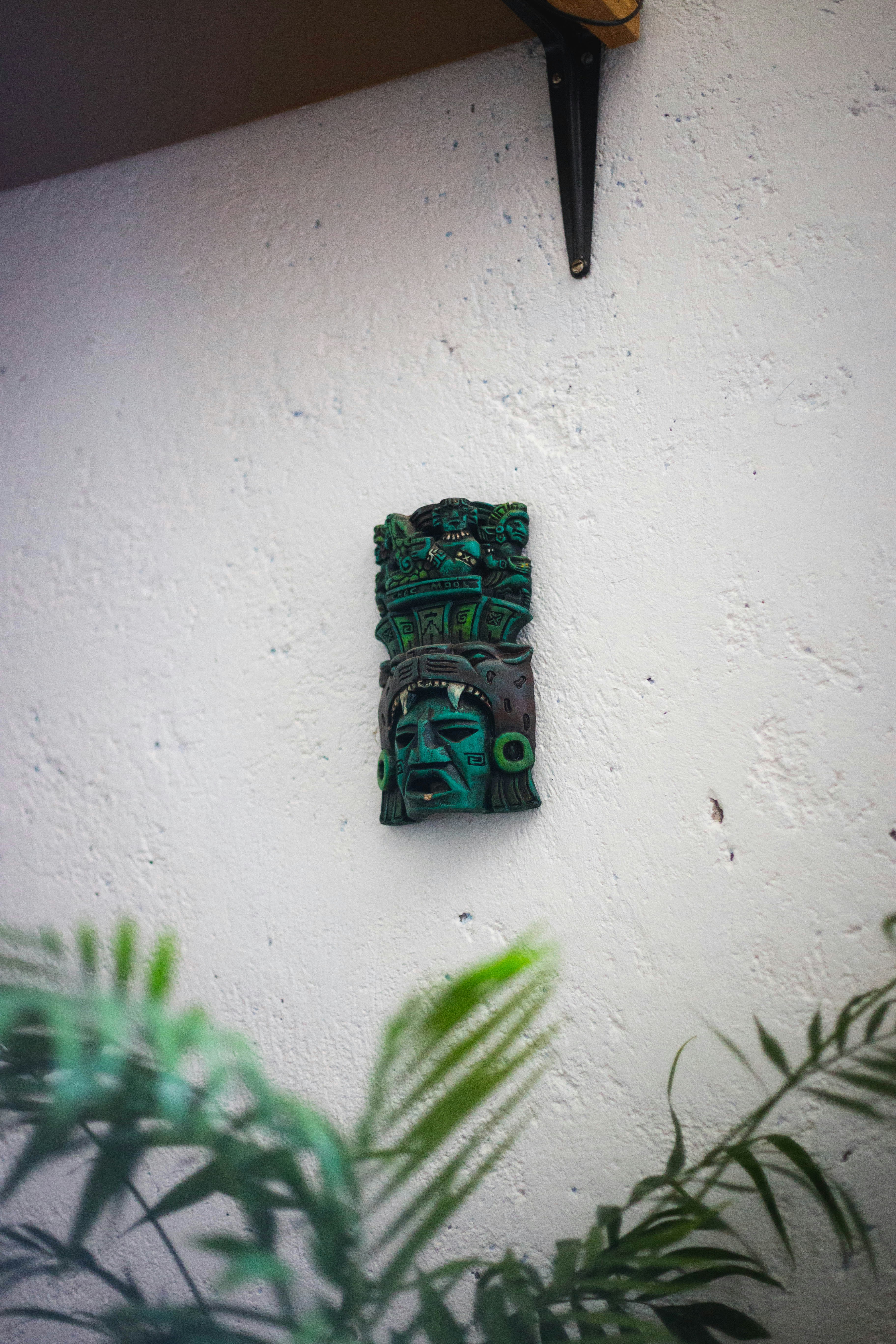 Intricately carved green mask mounted on a white wall, surrounded by lush foliage. The artwork reflects a rich cultural heritage.