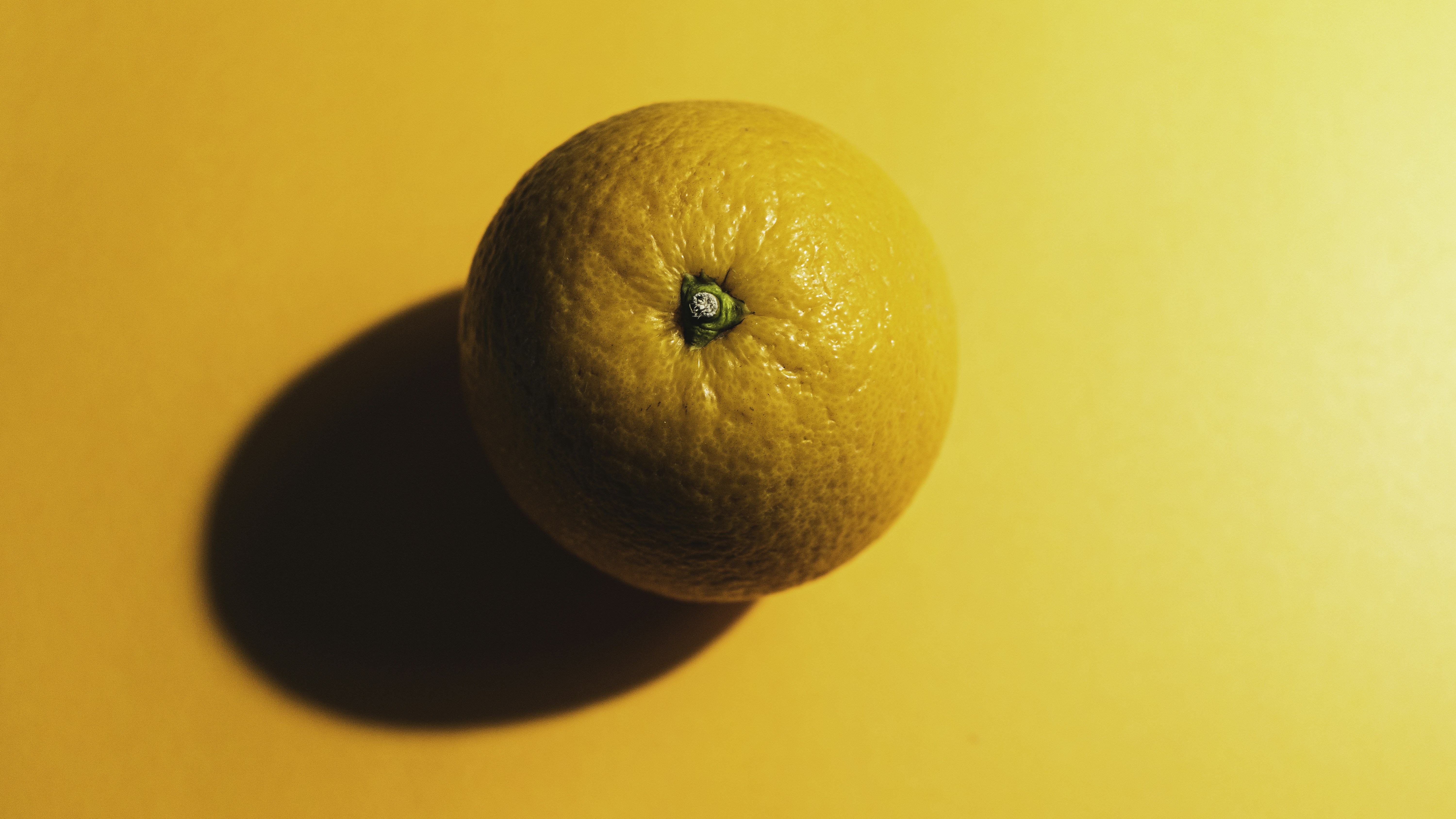 Vibrant yellow-orange lemon resting on a matching background, casting a soft shadow. The simplicity highlights the fruit's texture and color.