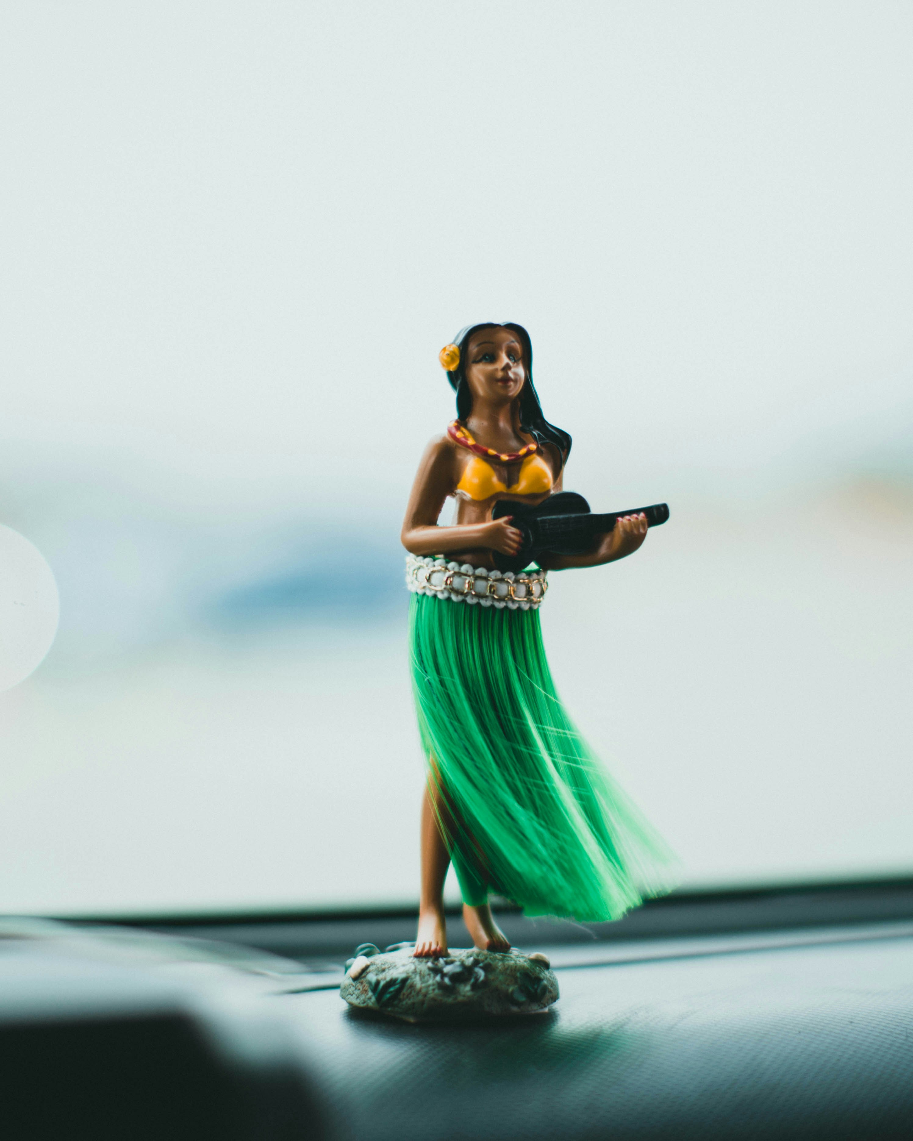 Hula dancer figurine adorned in traditional attire, strumming a ukulele, set against a softly blurred background.