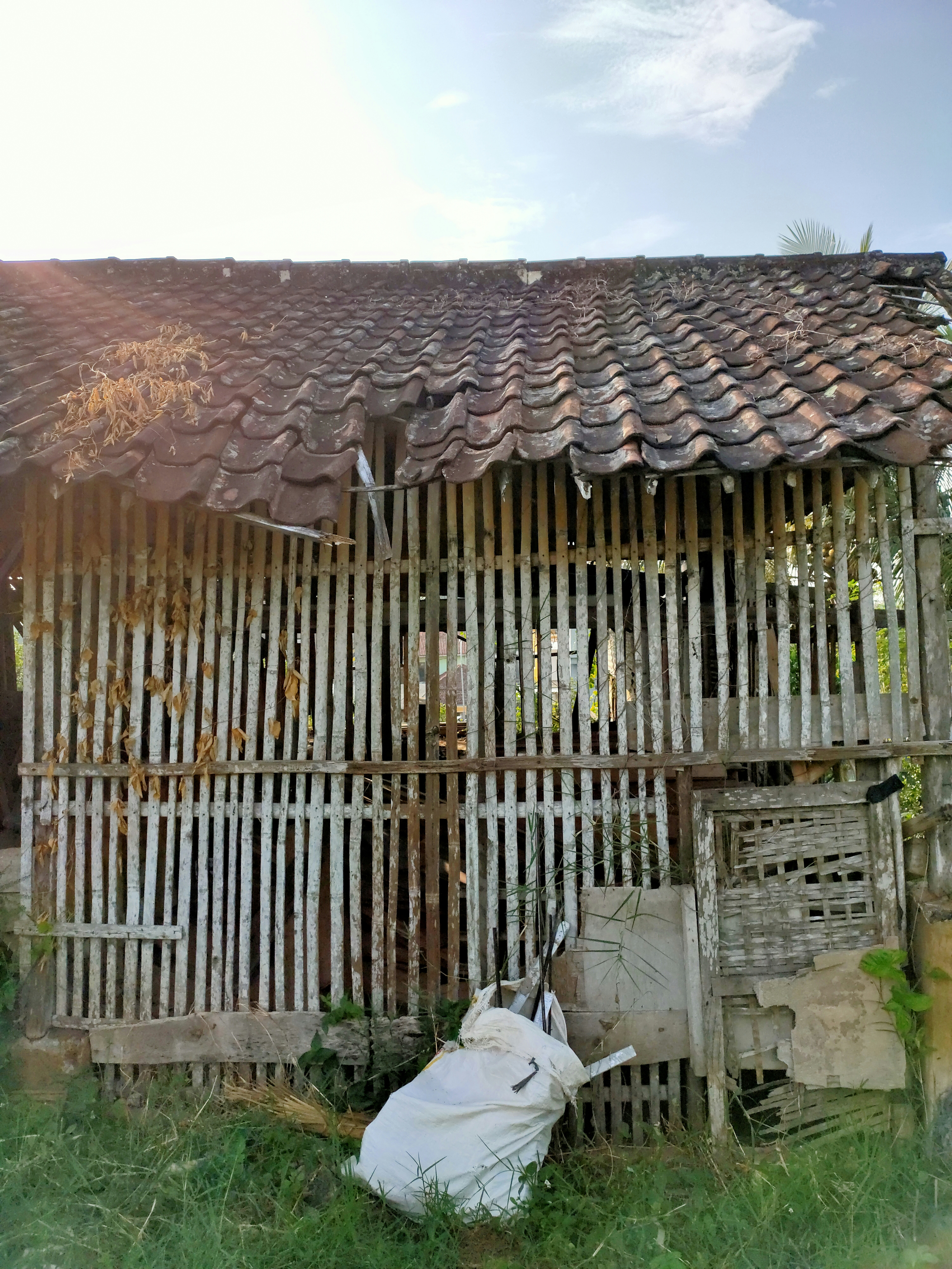 Weathered slatted shack with a tiled roof and a white bag in the foreground. Sunlit, rural scene captured in daylight.