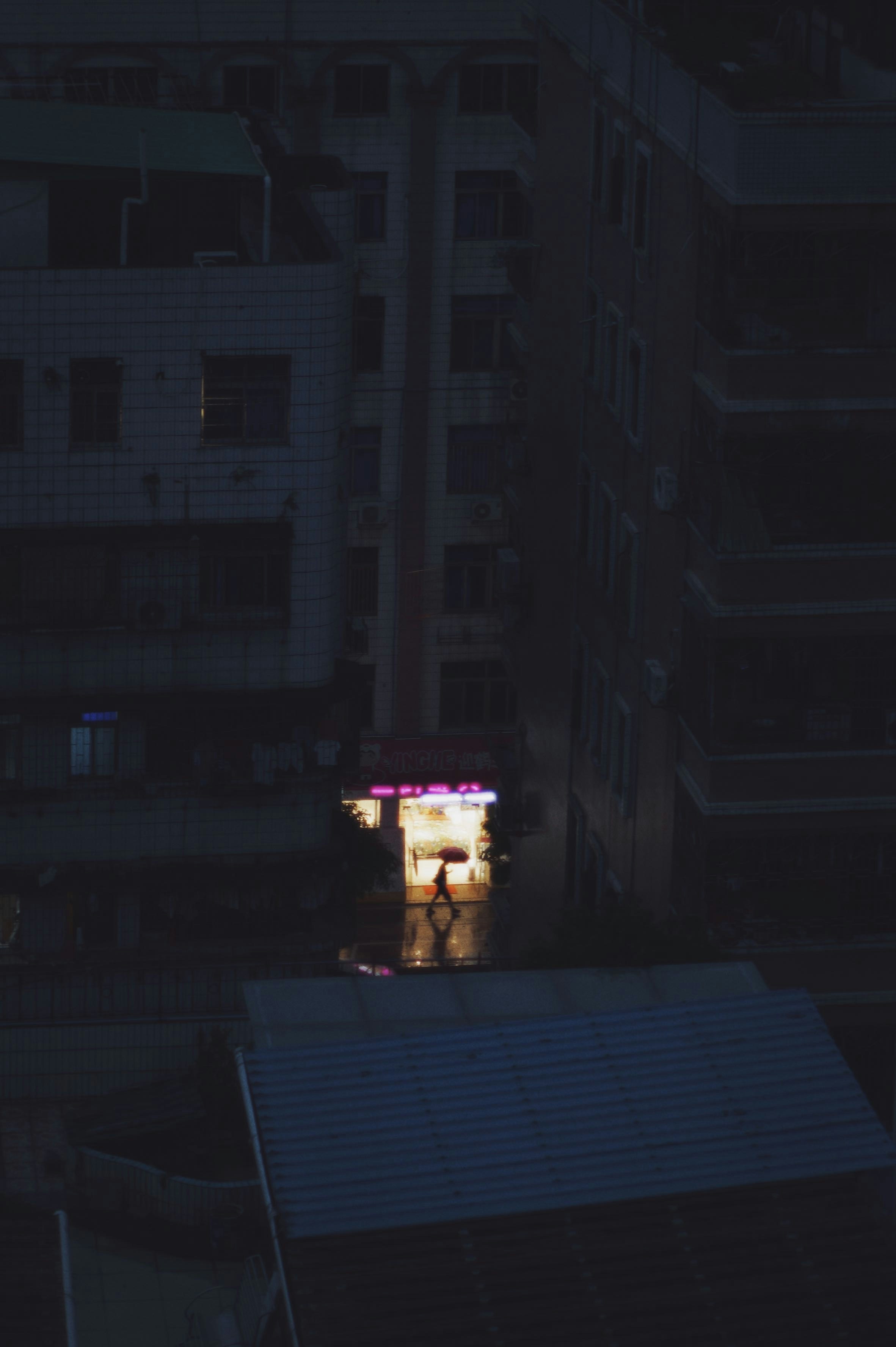 A lone figure walks past a brightly lit storefront amidst darkened buildings, highlighting the contrast between light and shadow in an urban setting.