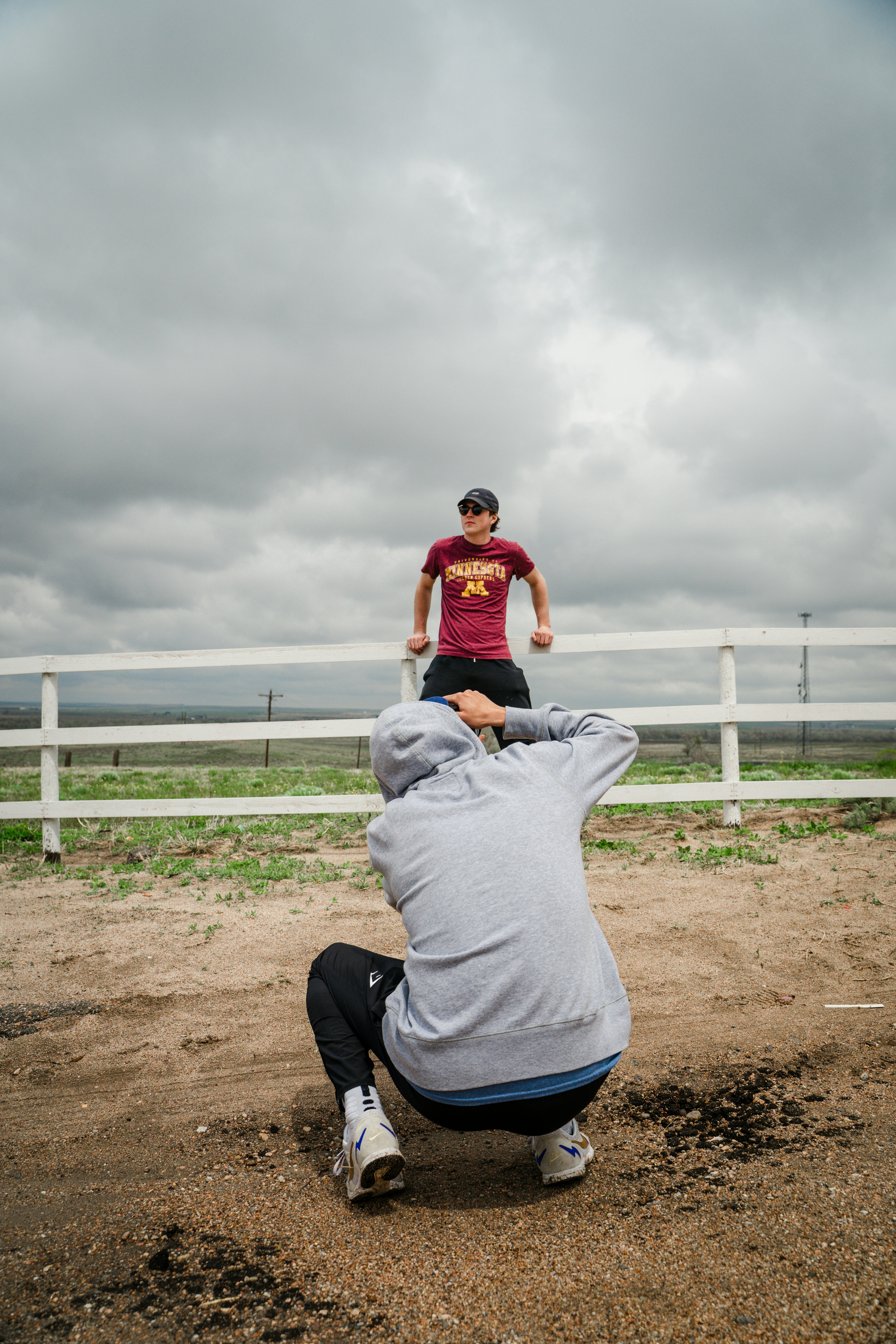 Man taking picture of other man in red shirt against white fence on a cloudy day