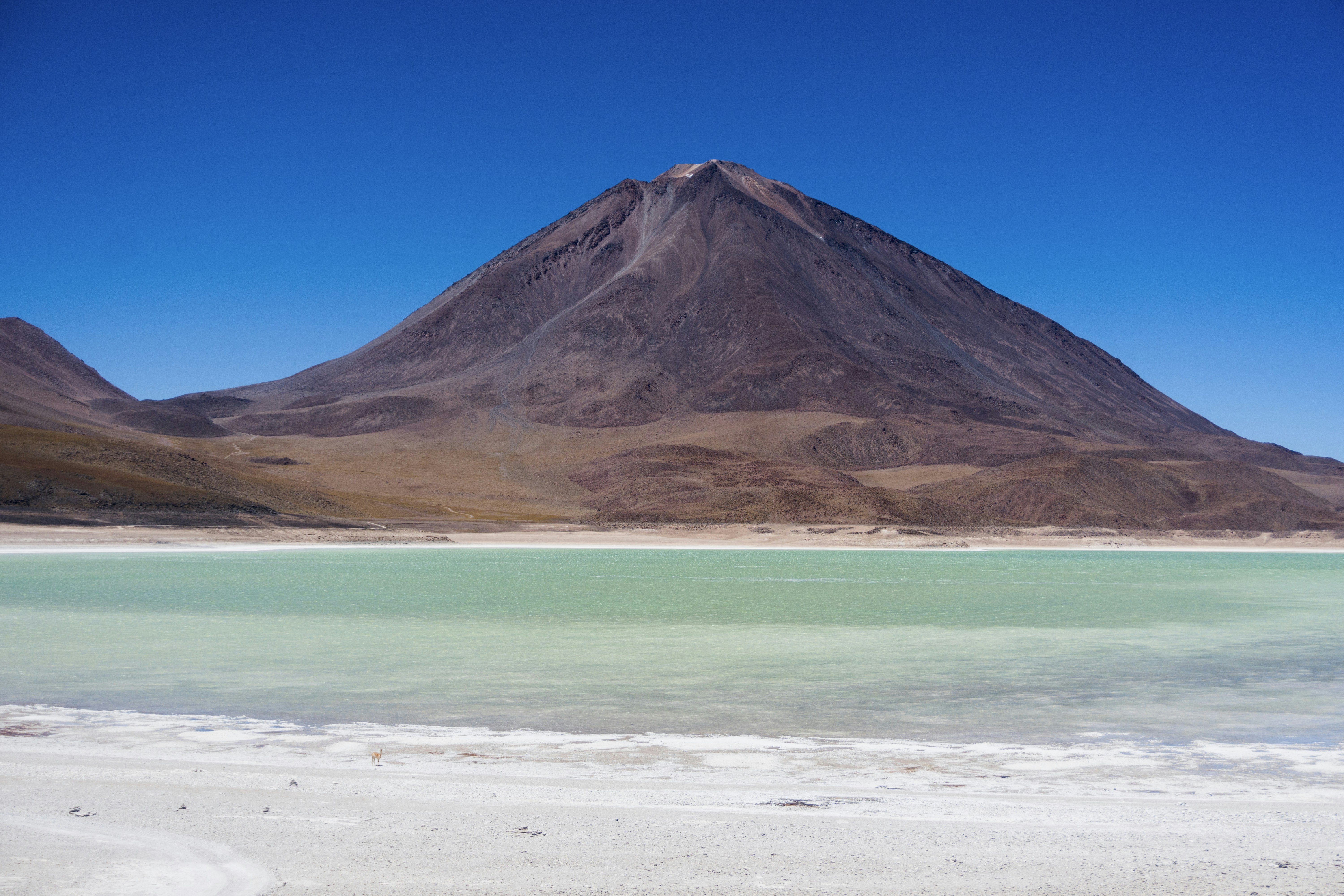 Turquoise waters of a high-altitude lake beneath a towering volcanic mountain under a clear blue sky.