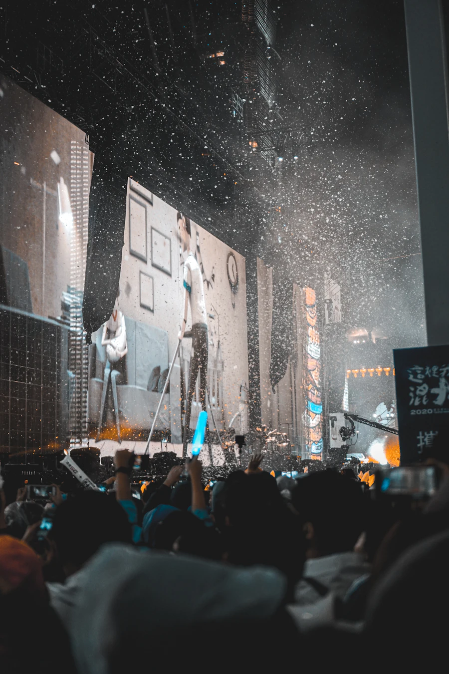 Crowded nighttime concert scene with a giant stage screen, falling confetti, and glowing signs in the background. Audience members raise phones and light sticks toward the performance.