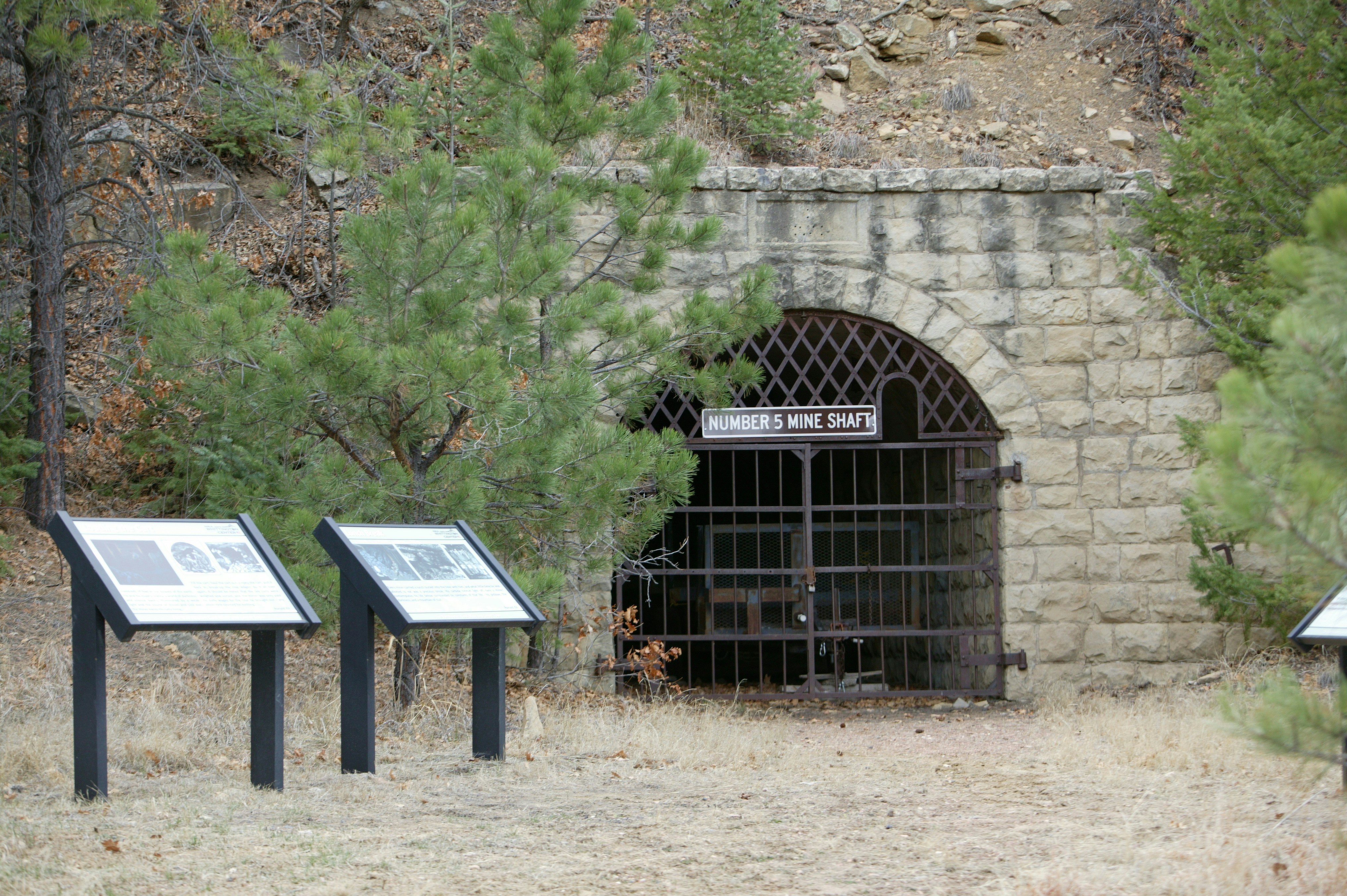 Historic mine entrance framed by pine trees, with informational signs nearby detailing its significance.