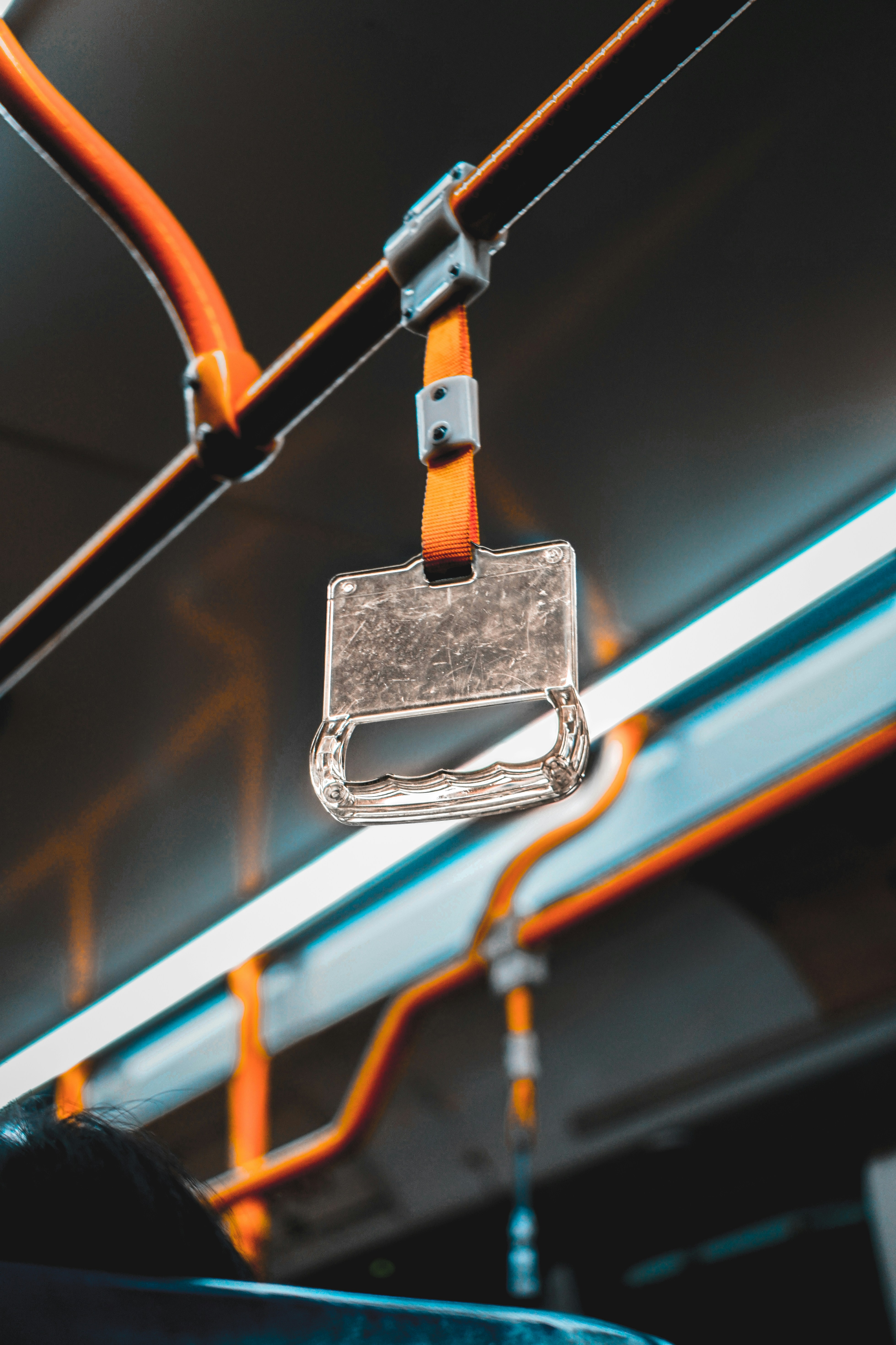 Metal hand strap hanging from a bus ceiling, illuminated by ambient light. The vibrant orange straps contrast against a sleek, modern interior.