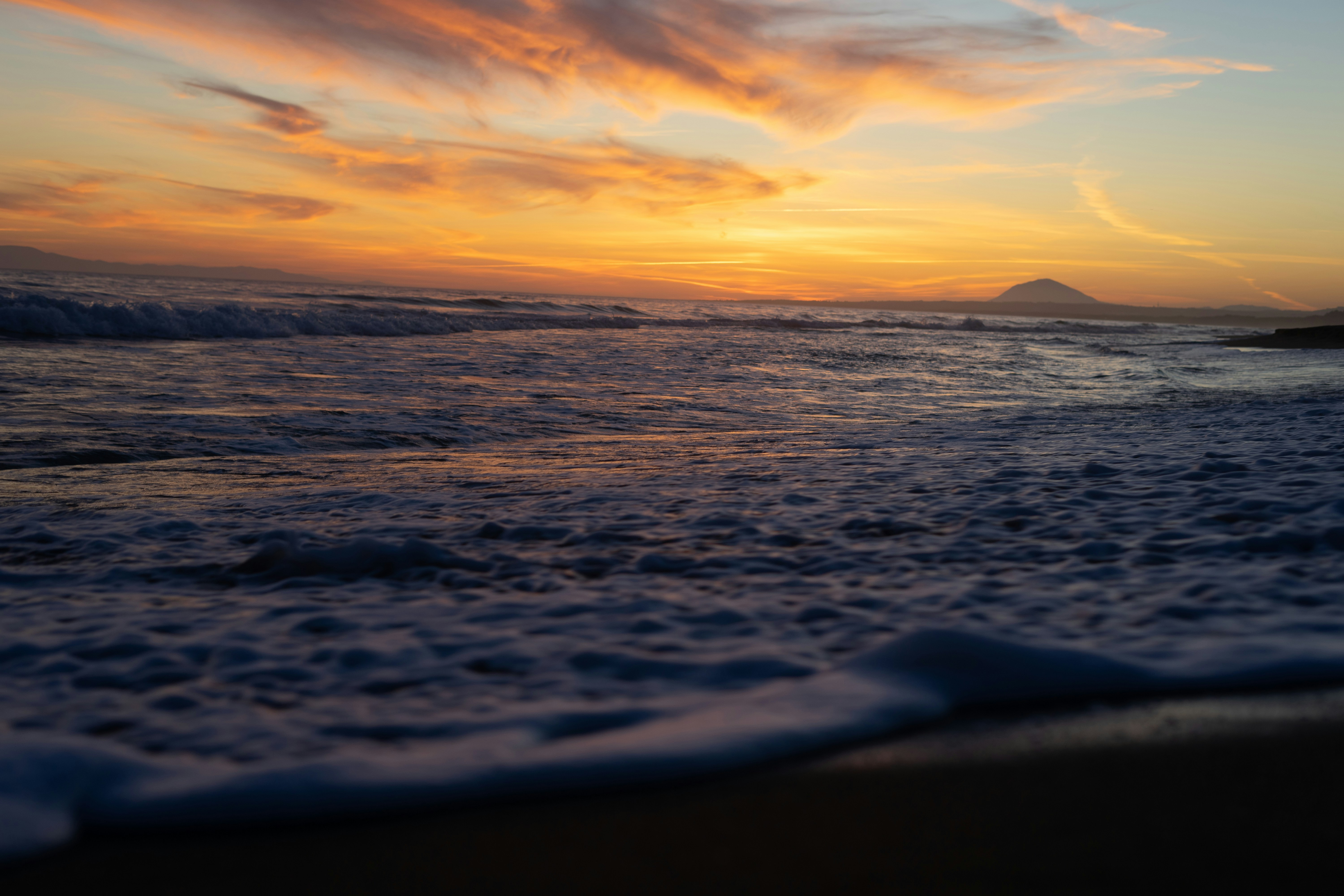 Gentle waves caress the shore as the sun sets behind distant mountains, casting warm hues across the sky. The scene captures a serene moment at the beach.