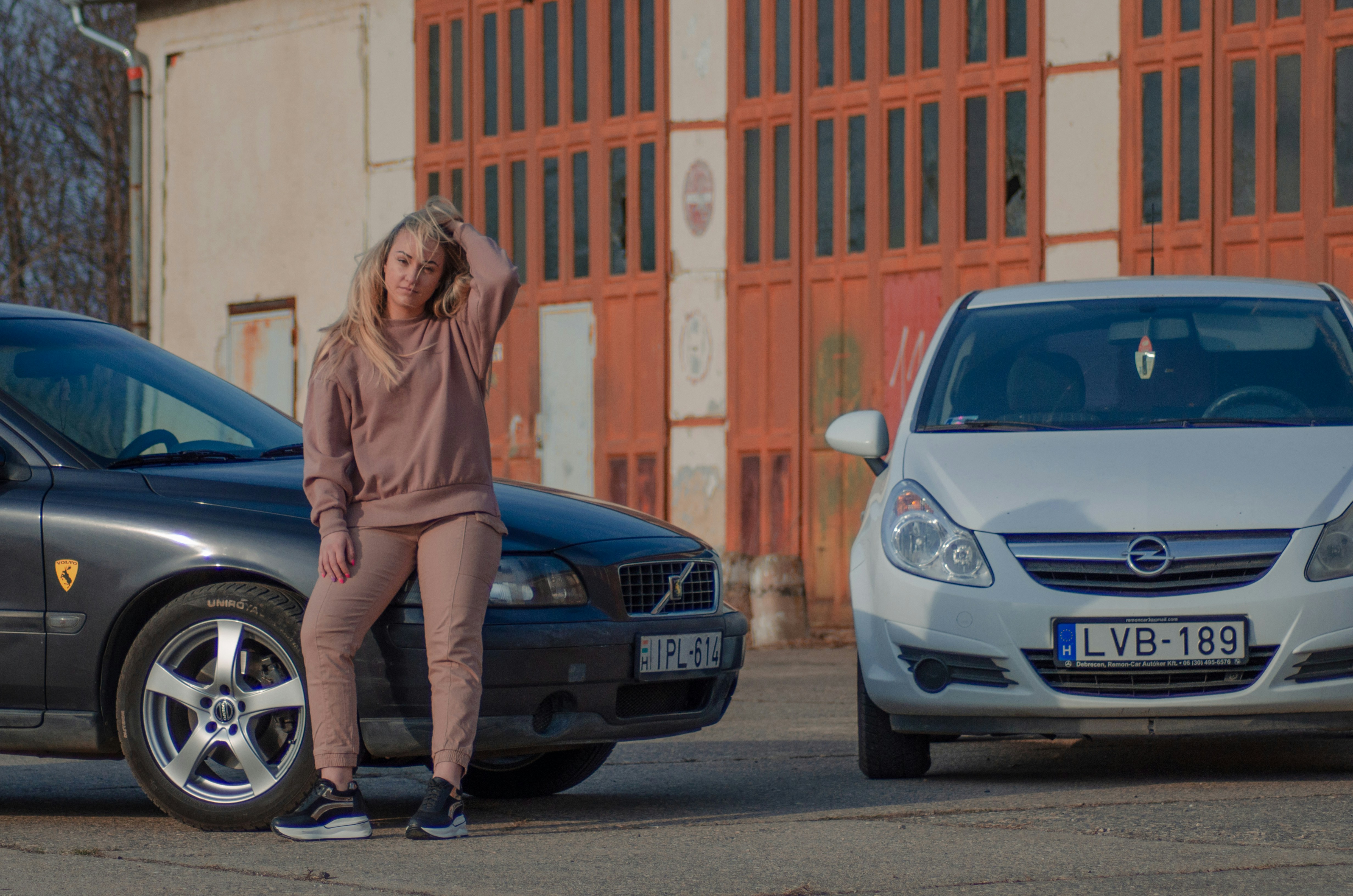 Person in casual wear leans against a black car with a white car nearby, in front of a vintage garage setting.