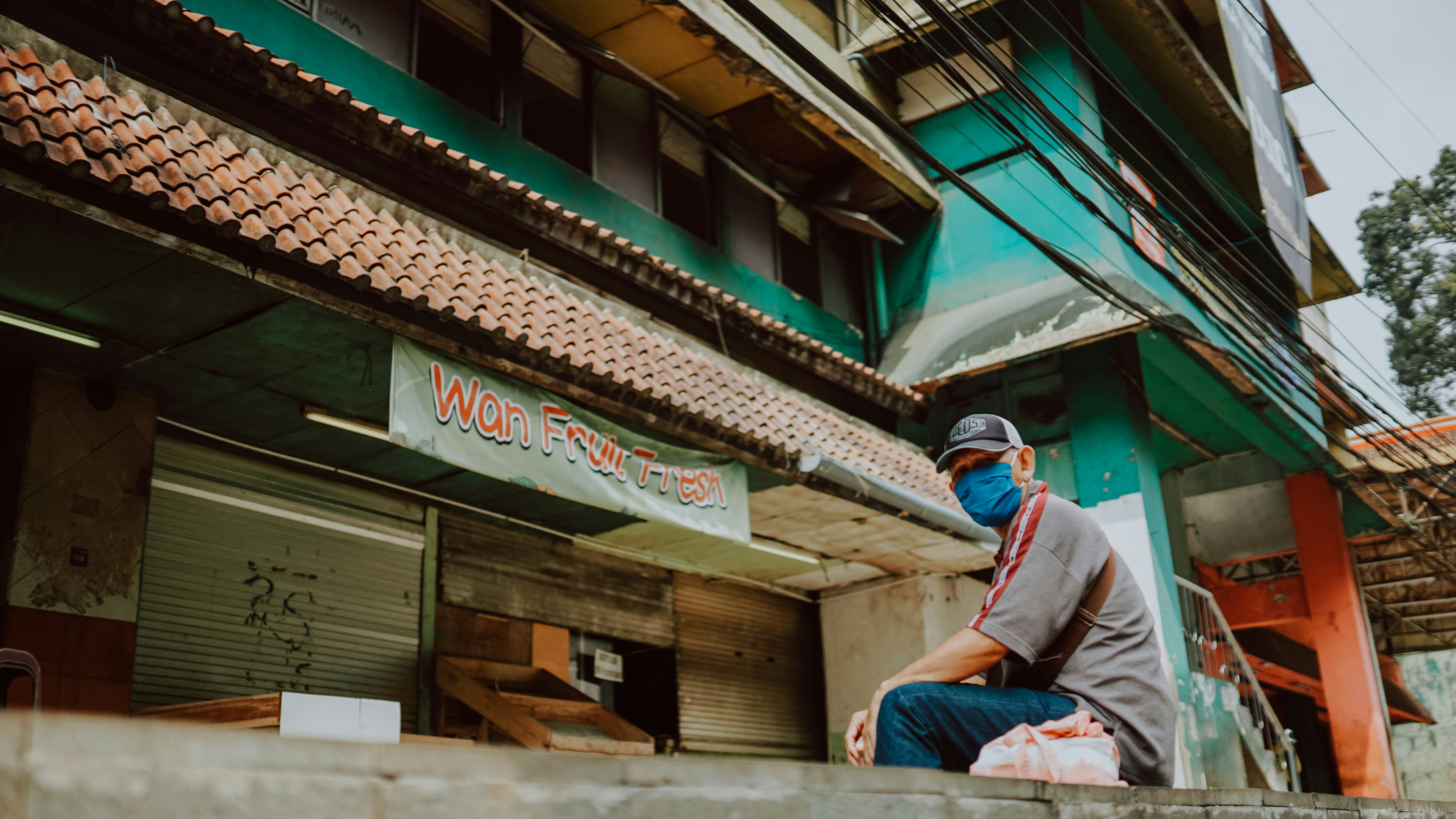 Man in blue jacket and denim jeans sitting on a blue plastic crate beside a shuttered storefront.