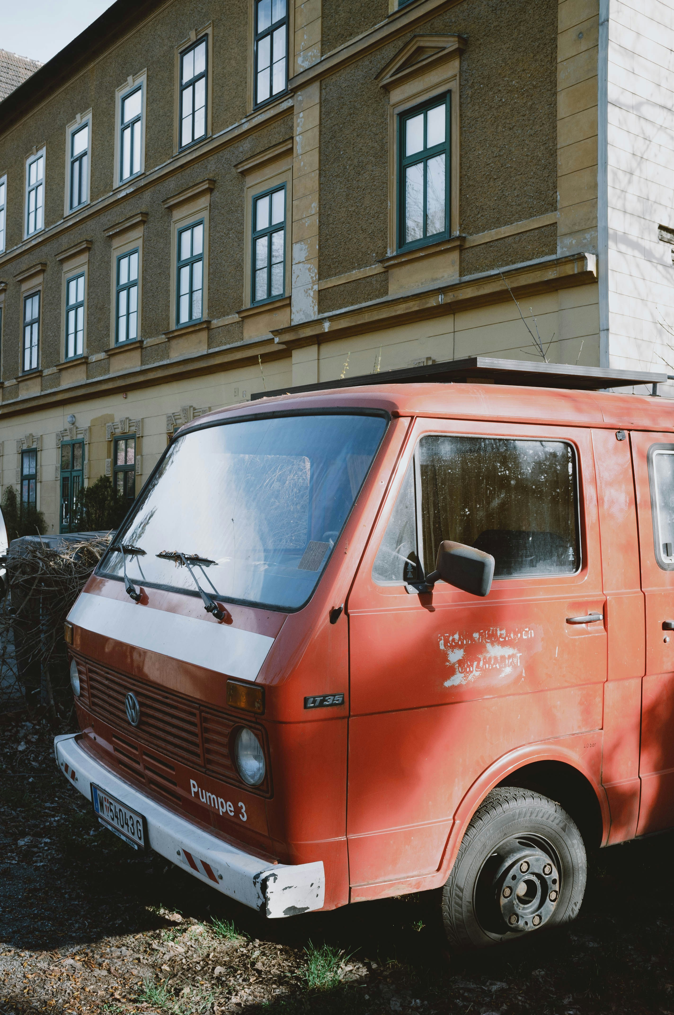 An abandoned red van with faded markings parked beside a historic building, showcasing a blend of nostalgia and decay.