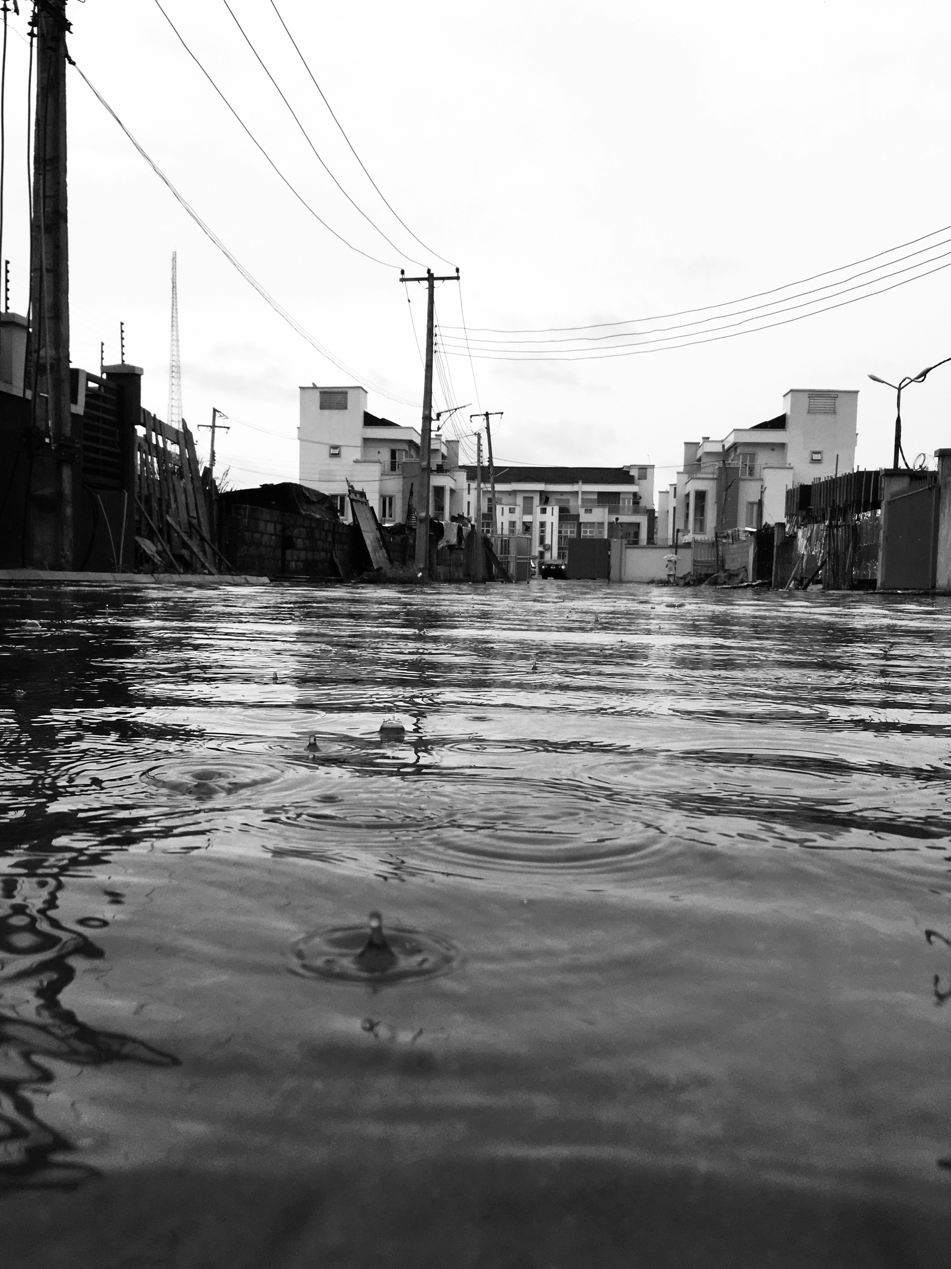 A flooded street reflecting the somber sky, with ripples forming on the water surface. Electrical poles and buildings line the background.