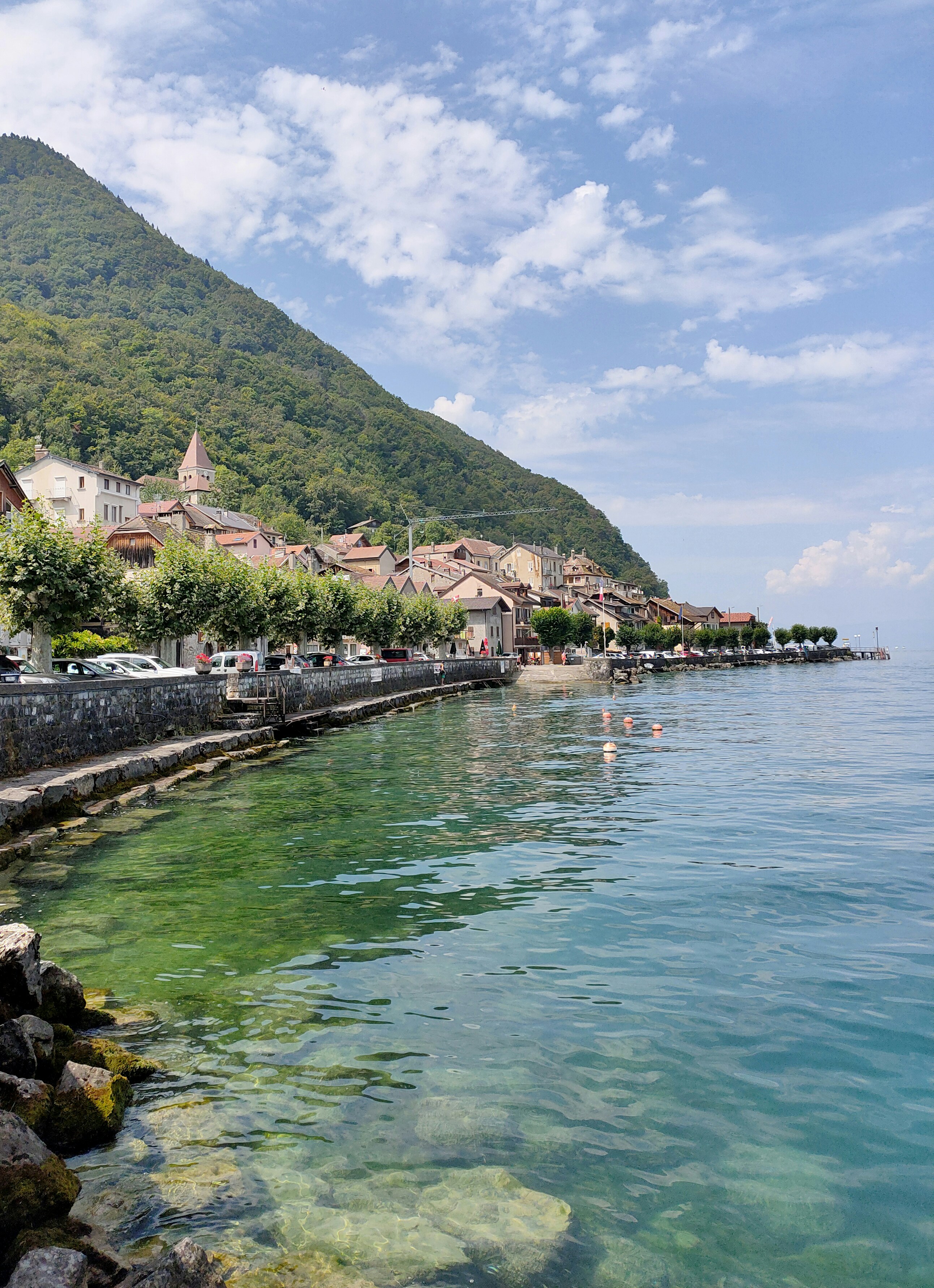 Coastal street with red-roofed houses along a stone quay, turquoise water, and a green mountain backdrop.
